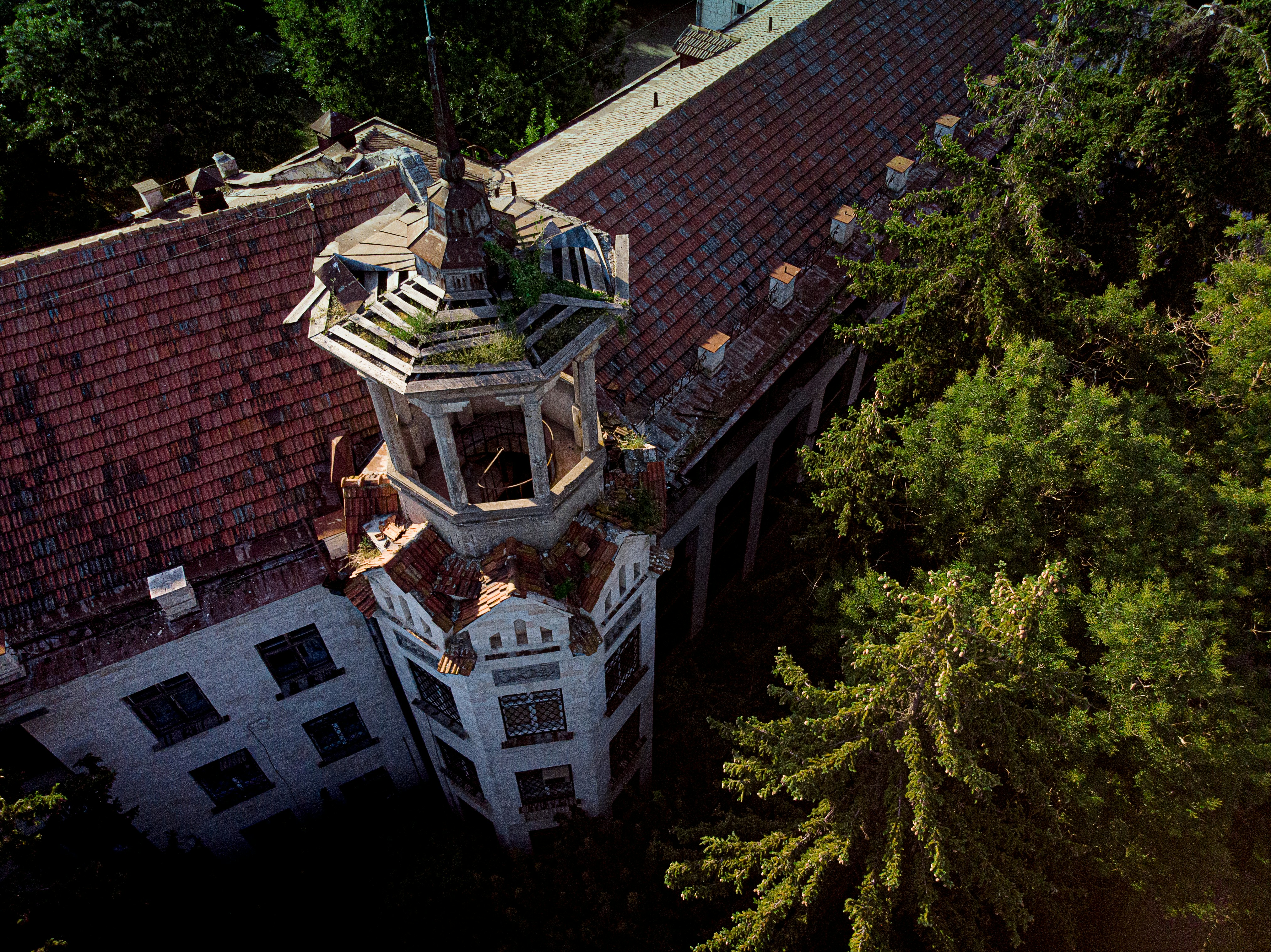 Aerial view of an abandoned building with a distinctive tower, surrounded by lush greenery. The weathered rooftop and architectural details hint at a rich history.