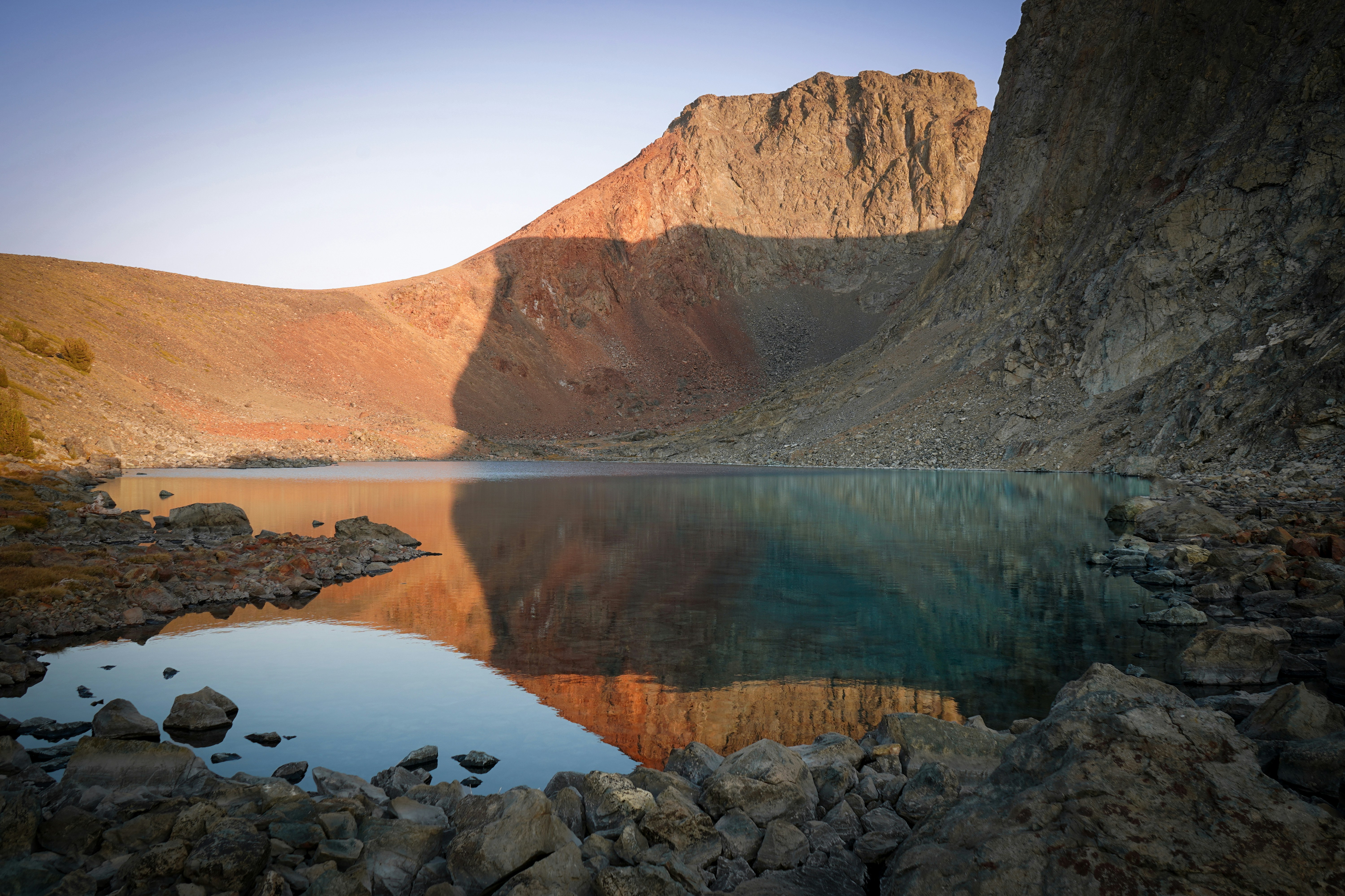 Tranquil alpine lake reflecting rugged mountain peaks as evening shadows stretch across rocky terrain.