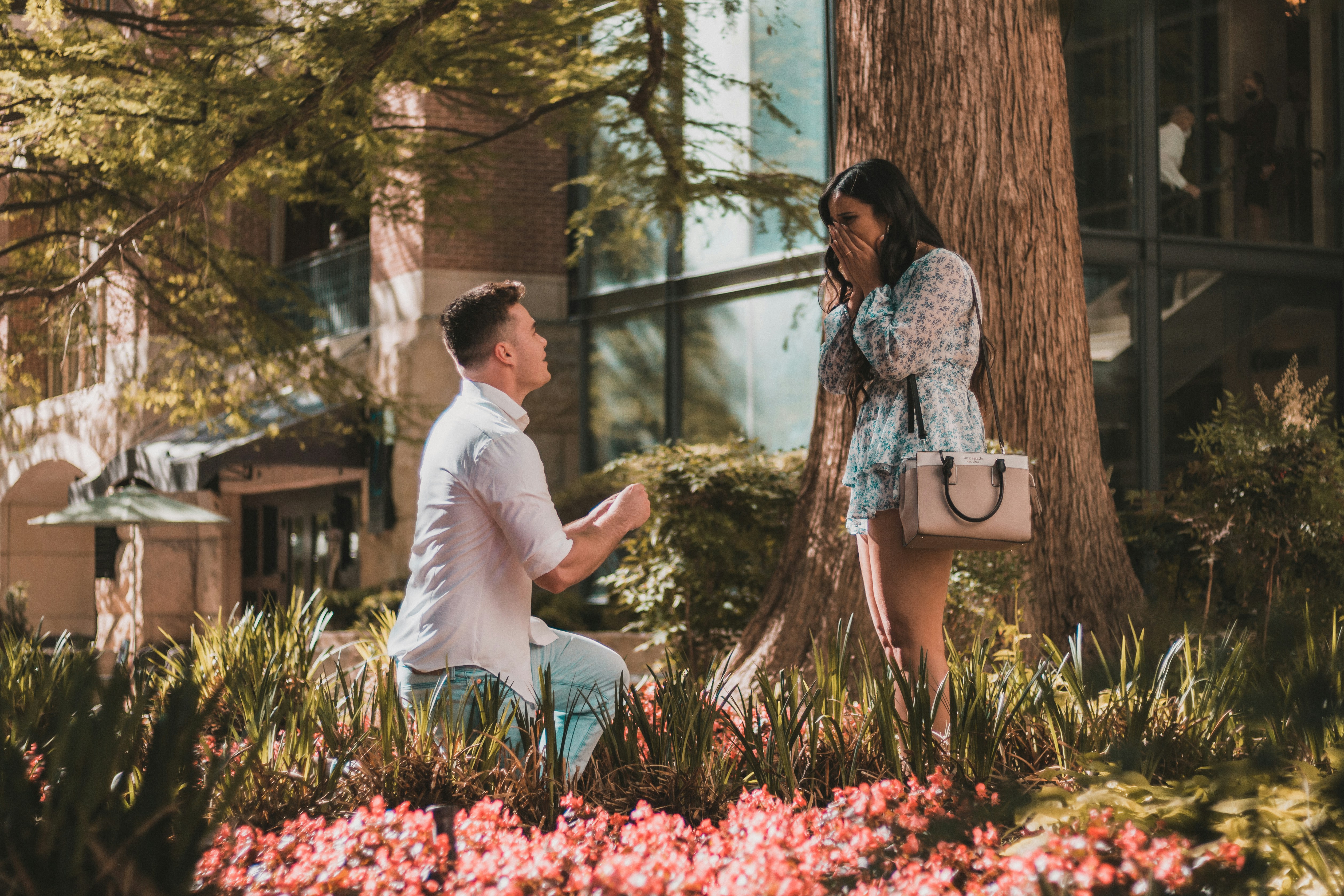 Couple in a garden as one person proposes on bended knee beneath a large tree.