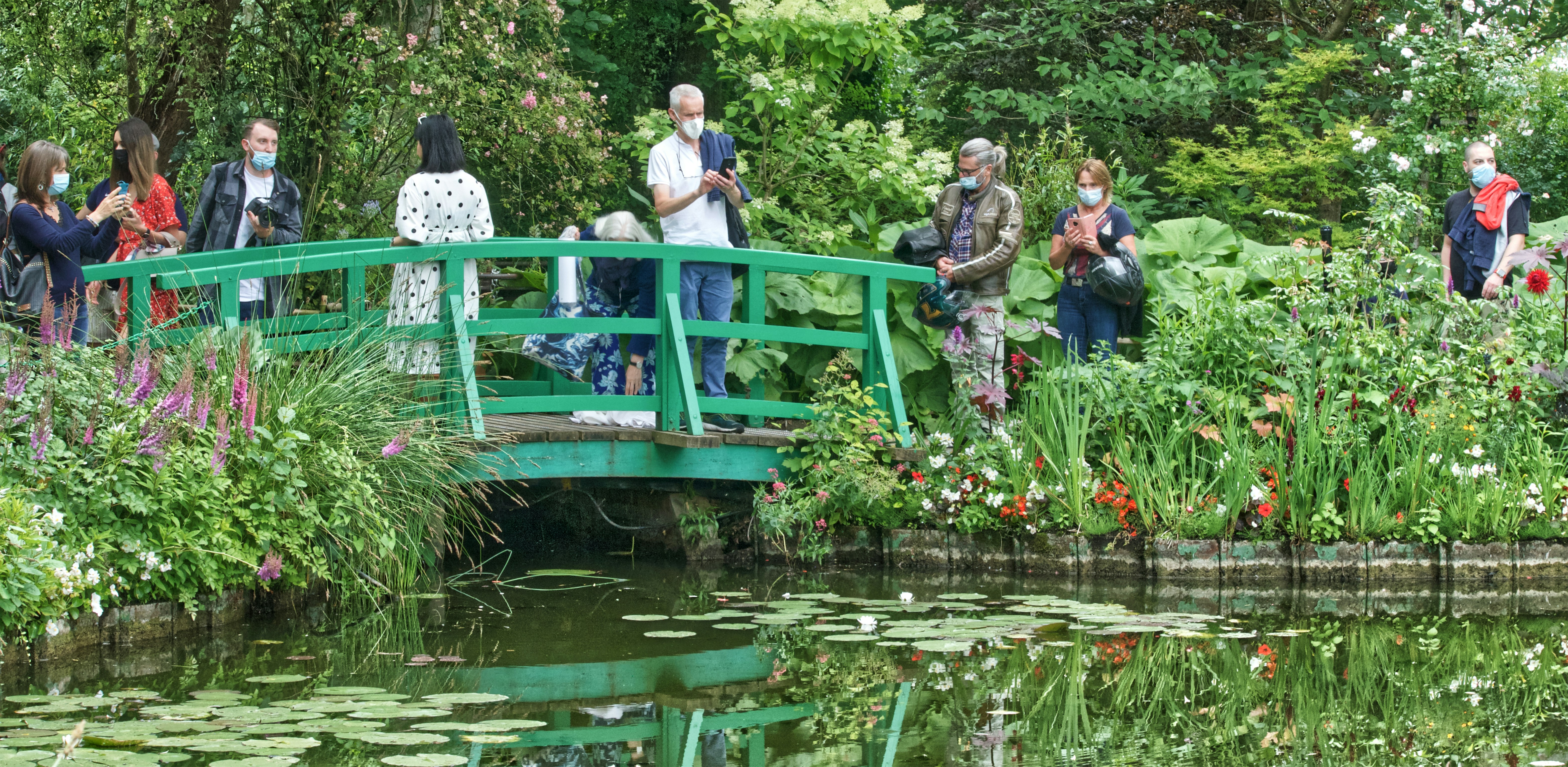 Visitors admire the lush garden surrounding a green wooden bridge, reflecting on the tranquil pond. The vibrant flora enhances the peaceful atmosphere.