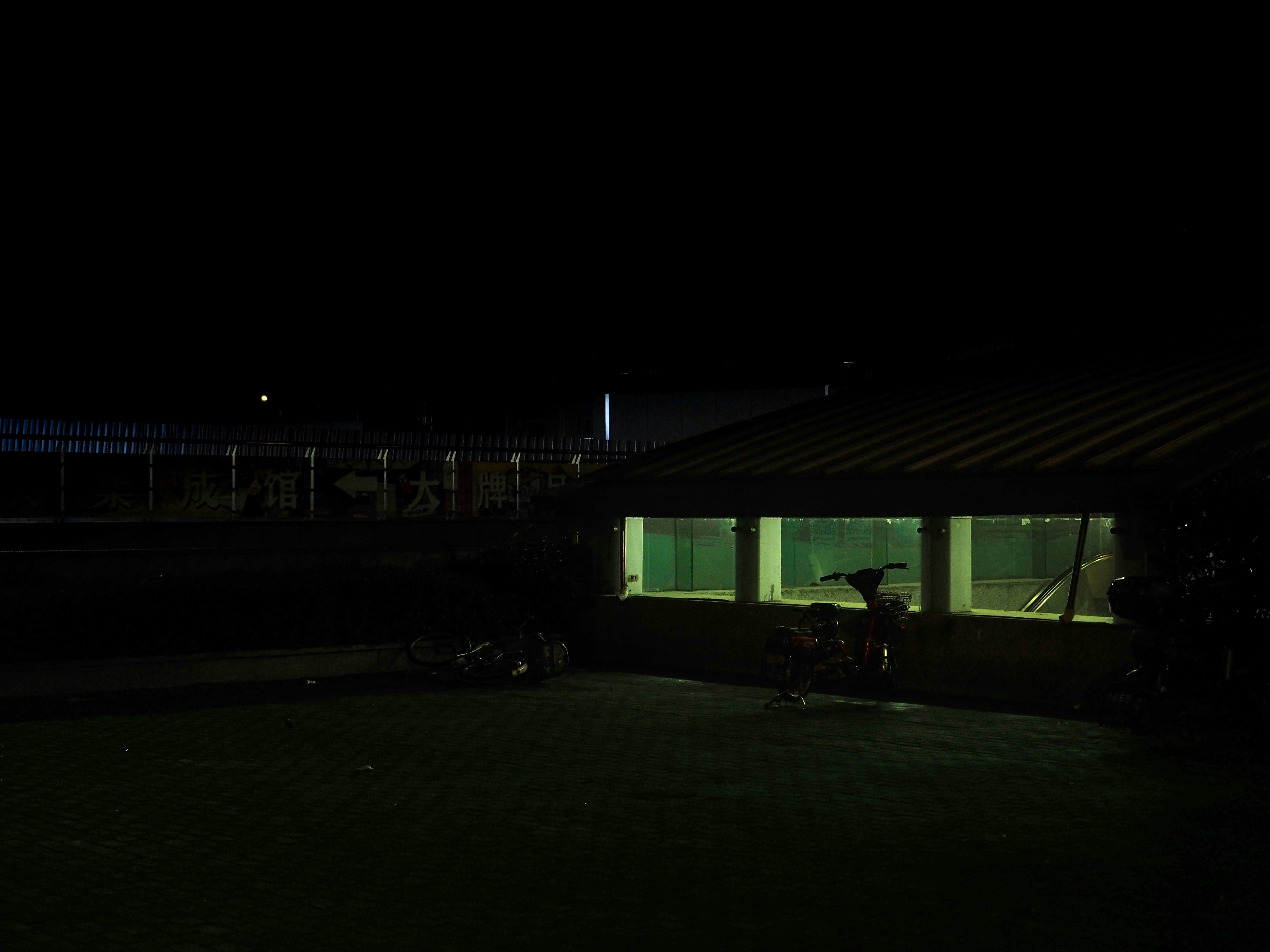 A dimly lit structure with green-tinted windows stands out against the night, surrounded by shadows and parked bicycles.