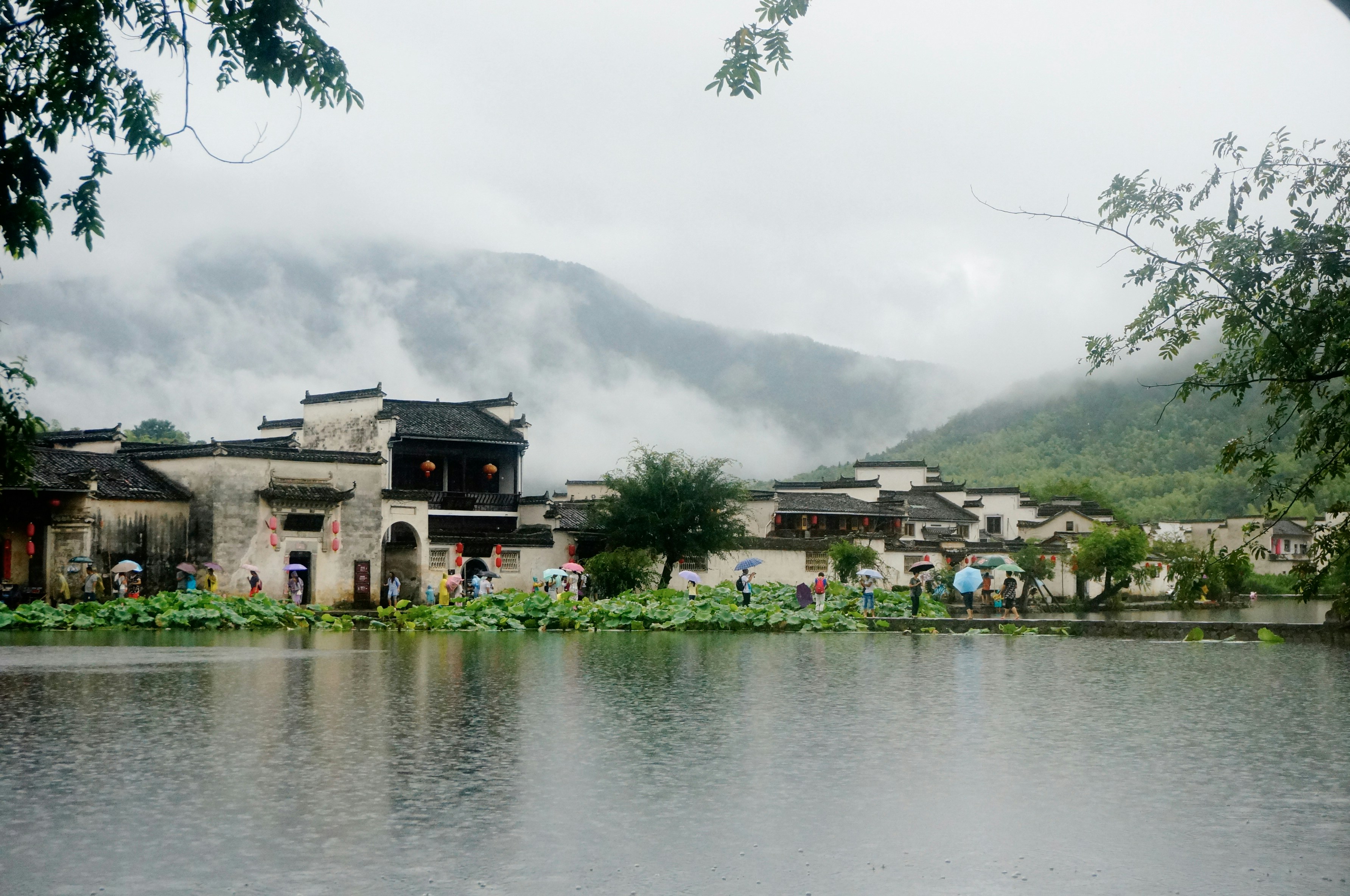 Traditional architecture lining a serene lake, shrouded in mist and rain, with colorful umbrellas dotting the scene.