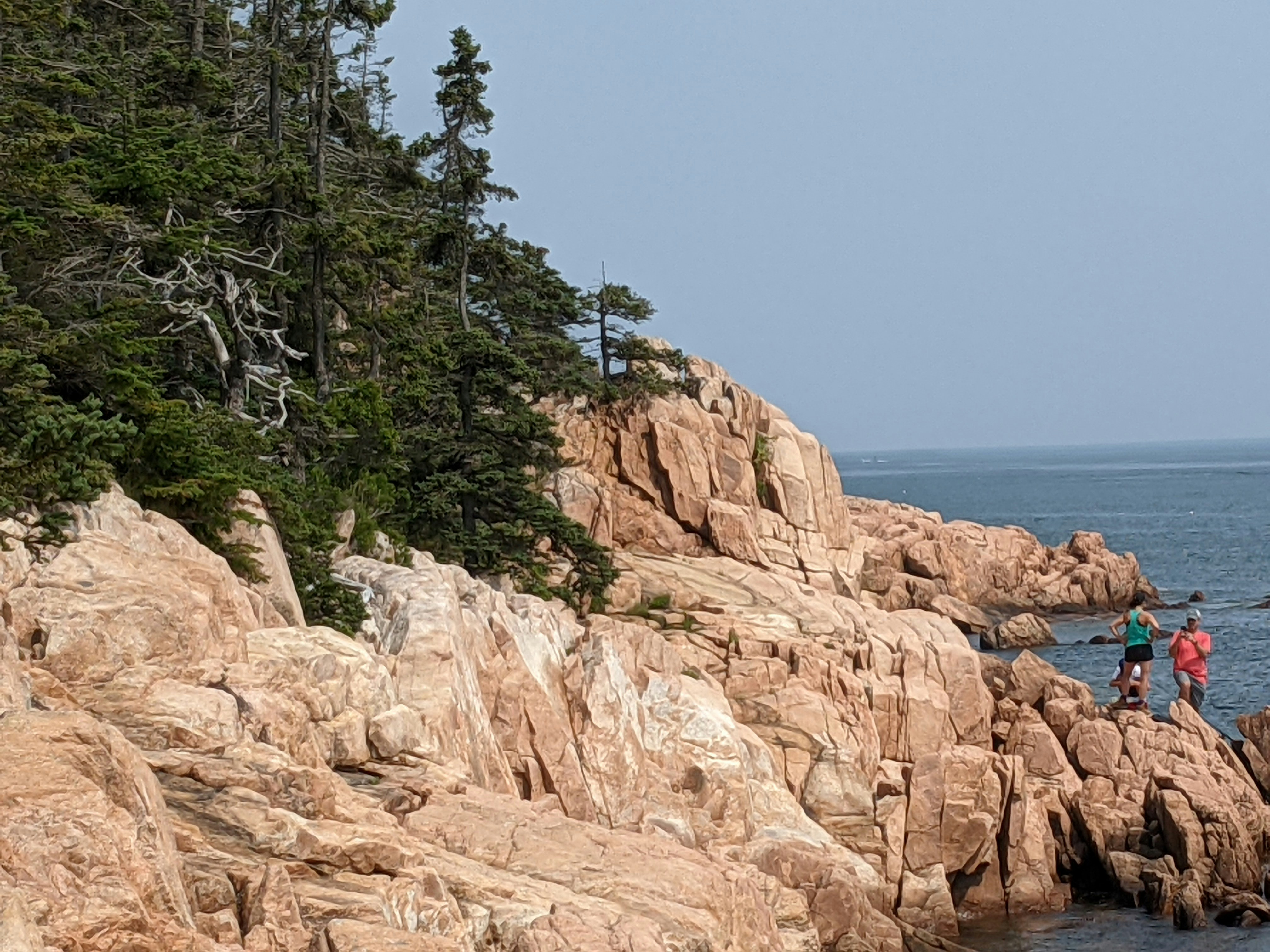 Two individuals standing on rocky shoreline, surrounded by lush greenery and the sea. The rugged landscape showcases the beauty of coastal terrain.