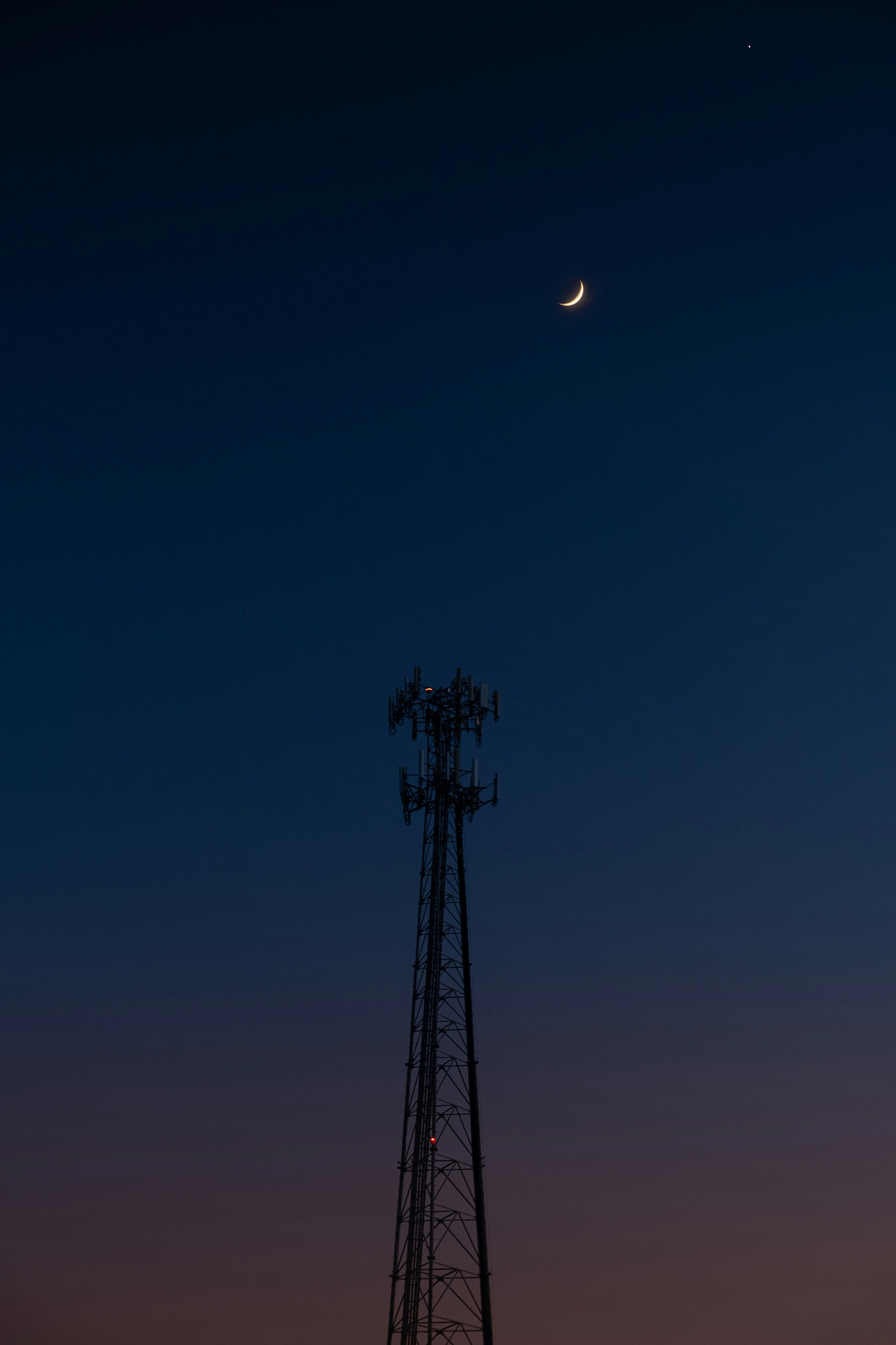 A telecommunications tower silhouetted against a twilight sky, with a crescent moon hanging above. Stars twinkle faintly in the deepening blue hue.