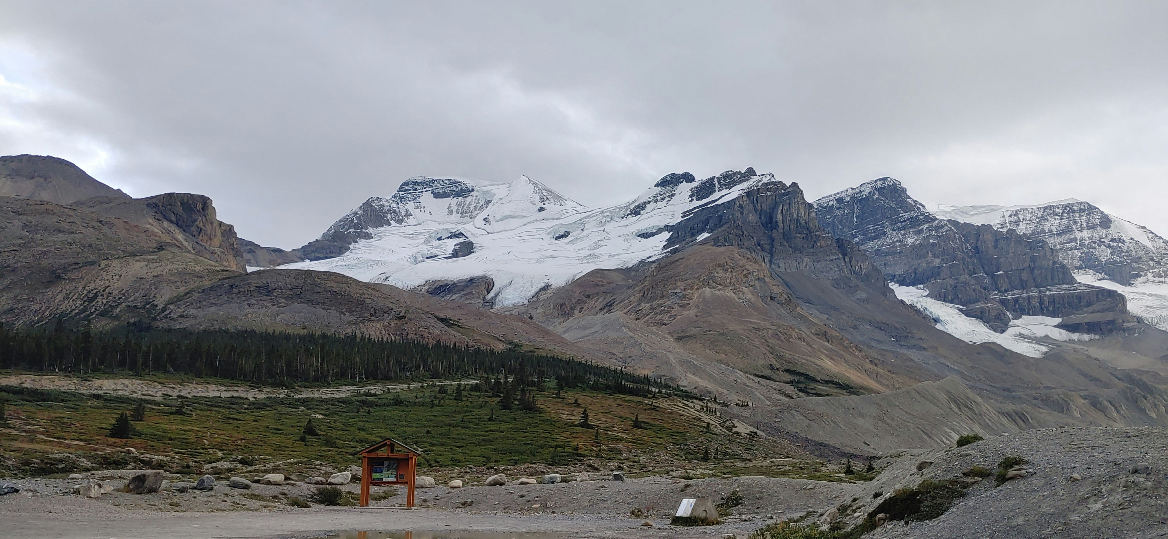 Snow-dusted alpine peaks rise above a rugged valley, with a small orange gate in the foreground beneath a heavy, overcast sky. The scene emphasizes scale, solitude, and the raw textures of rock and snow.