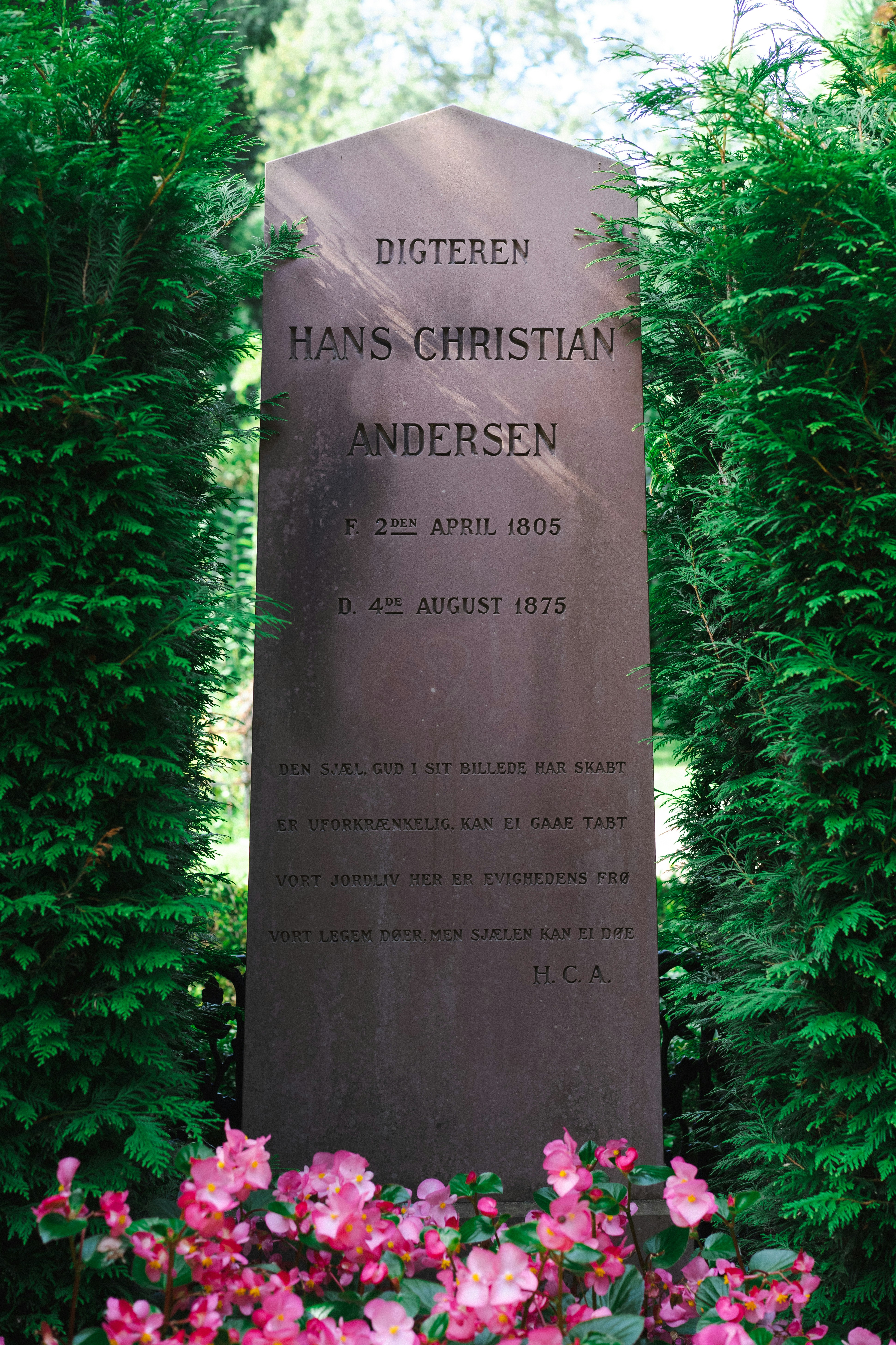 Gravestone of Hans Christian Andersen, surrounded by lush greenery and vibrant flowers, marking his birth and death dates. The inscription reflects his literary legacy.