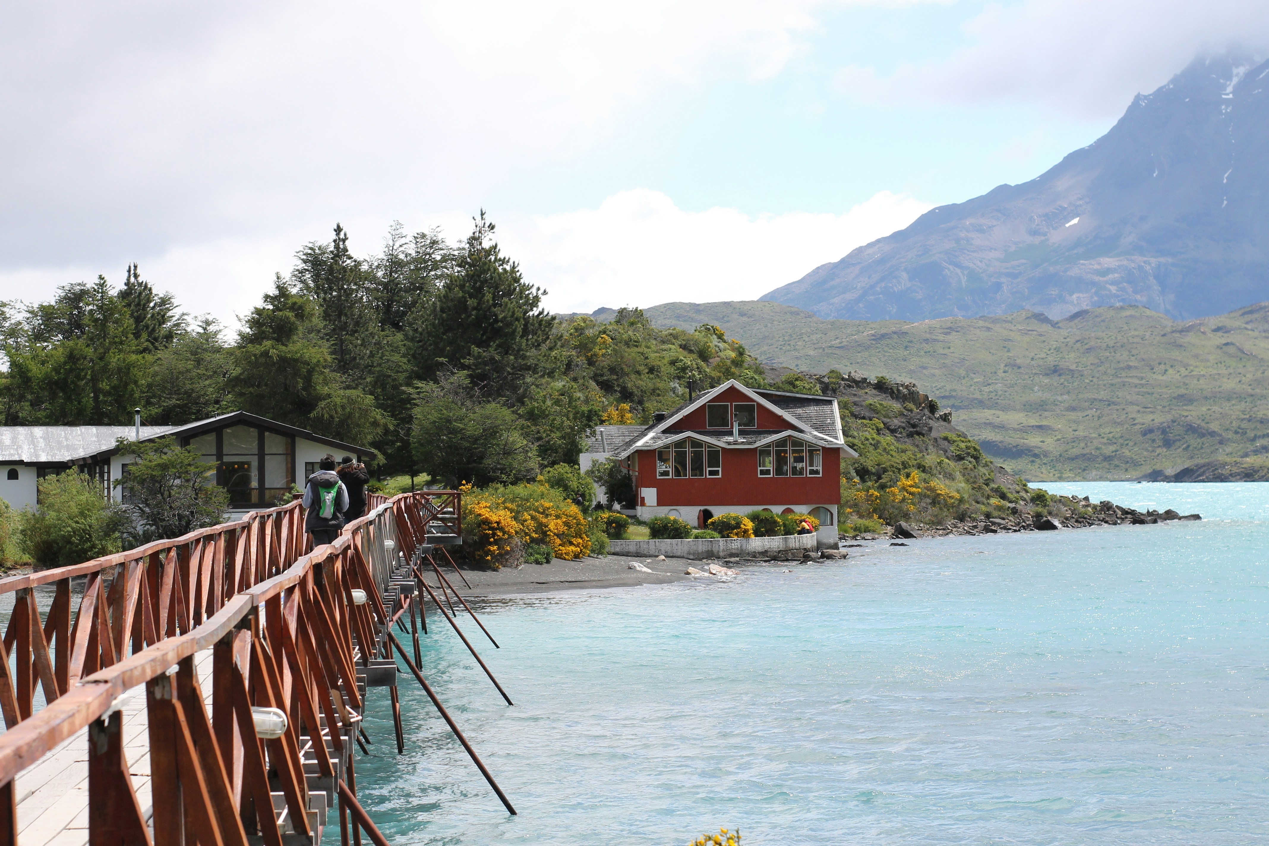 Red house nestled by a turquoise lake, with a bridge leading to it, surrounded by lush greenery and distant mountains.