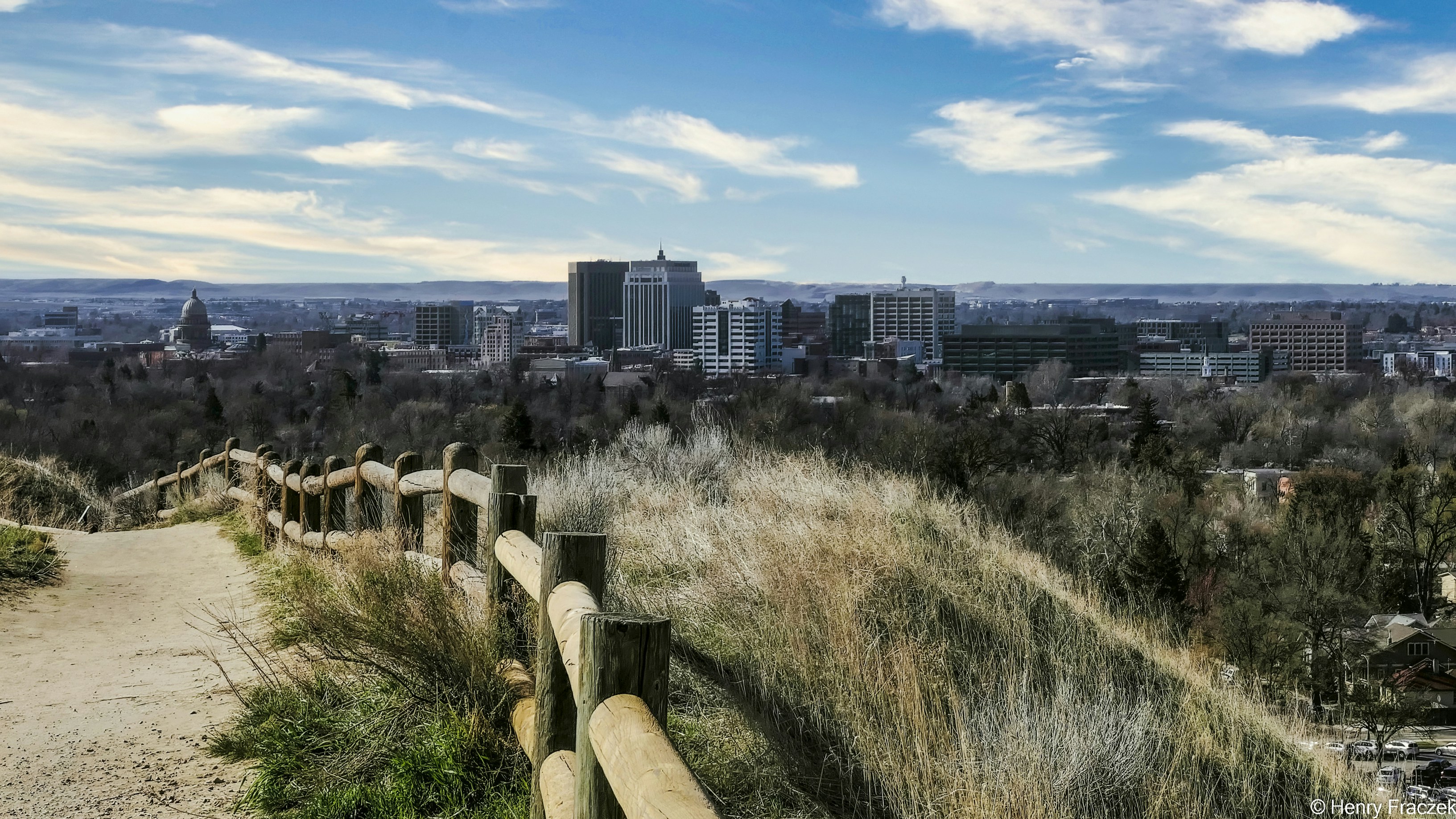 Rustic path with wooden fencing overlooks a city skyline under a vibrant blue sky.