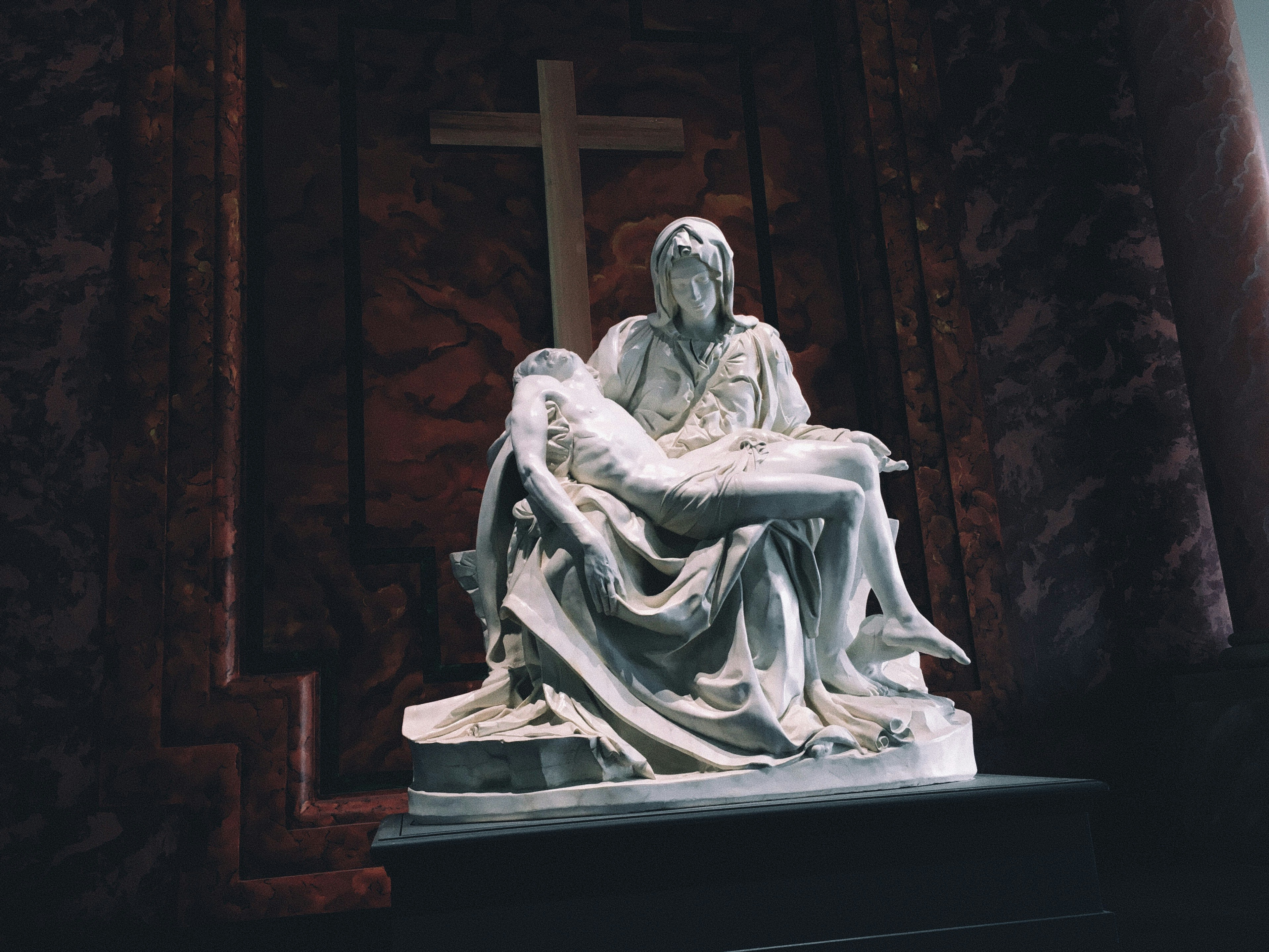 A marble Pieta-style sculpture cradled in a dim chapel, with a cross backdrop and draped fabric highlighted by soft, directional light.