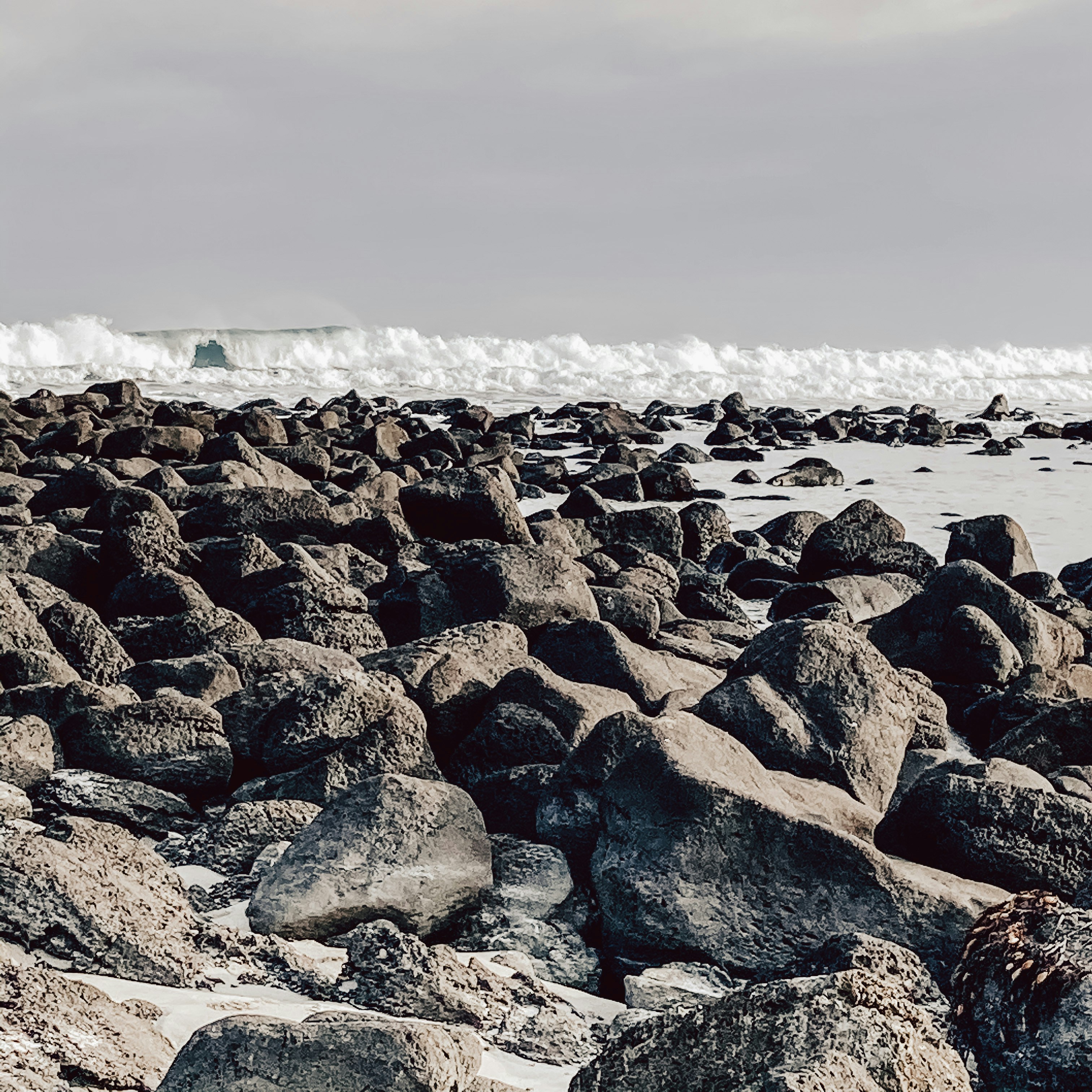 Jagged rocks scattered along a sandy beach, with waves crashing in the background under a cloudy sky.