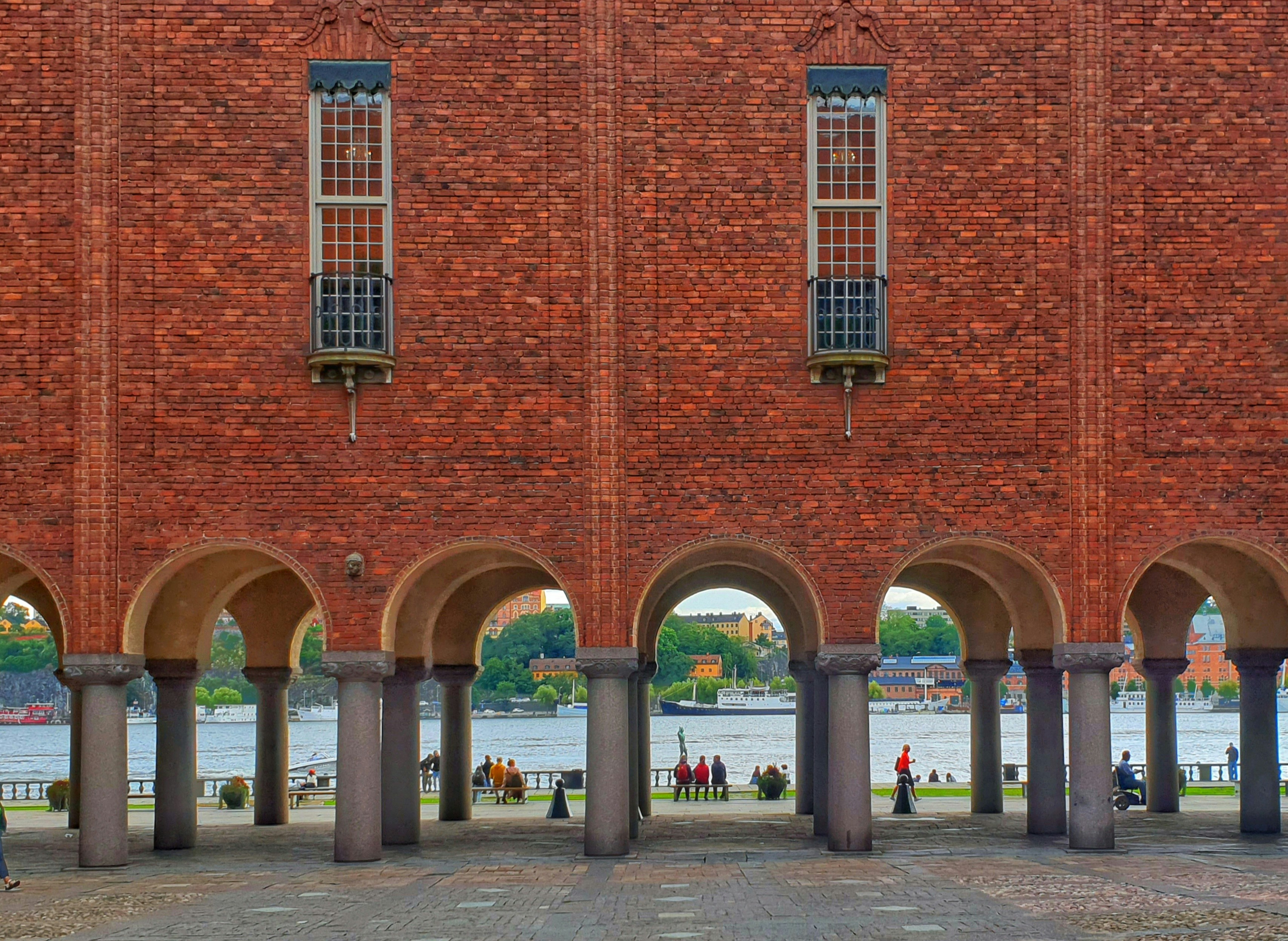 Historic brick building with arched columns framing a scenic waterfront view, showcasing people enjoying the space.