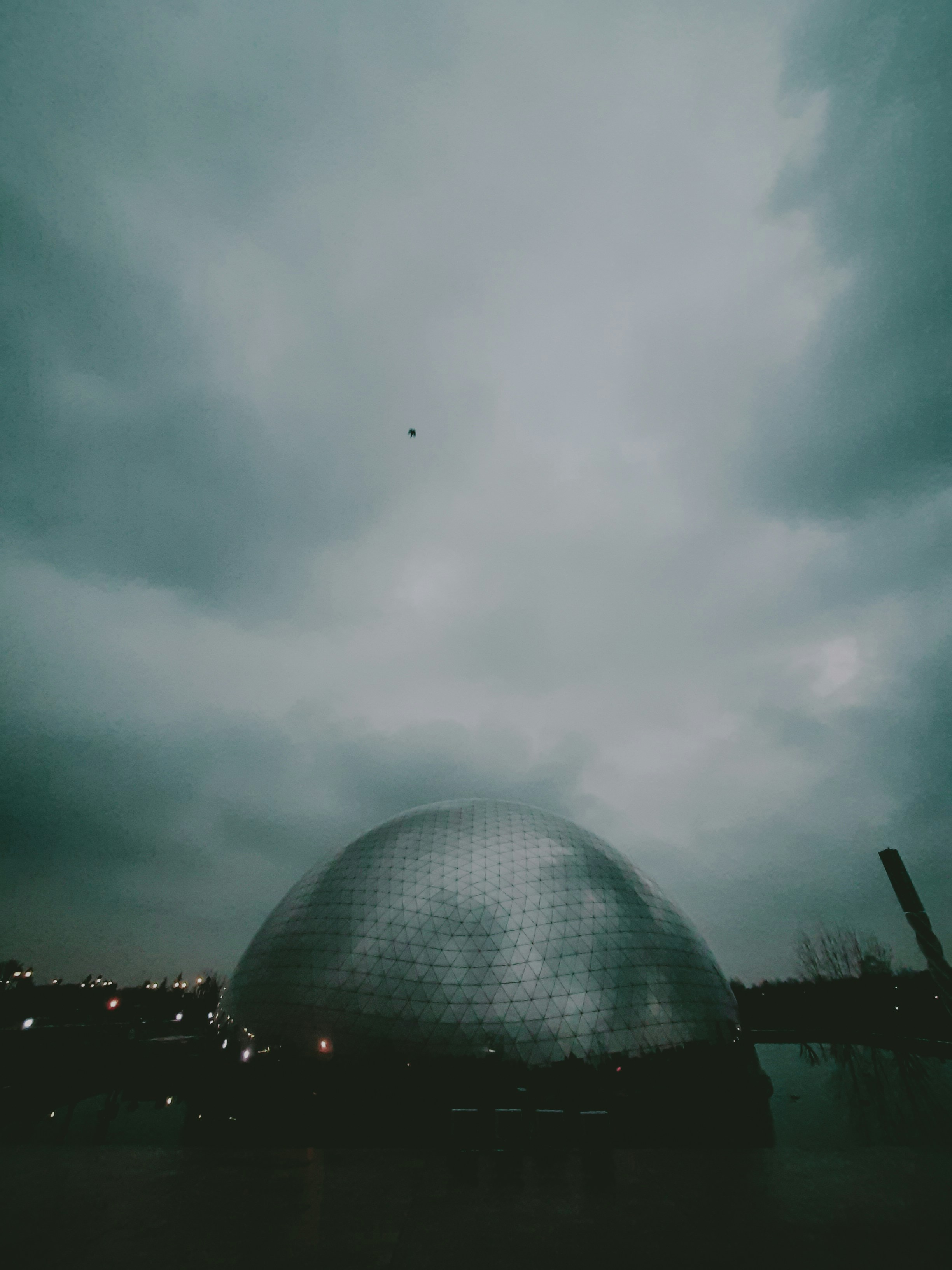 A large, transparent dome structure reflects the moody sky, surrounded by soft lights along the water's edge.
