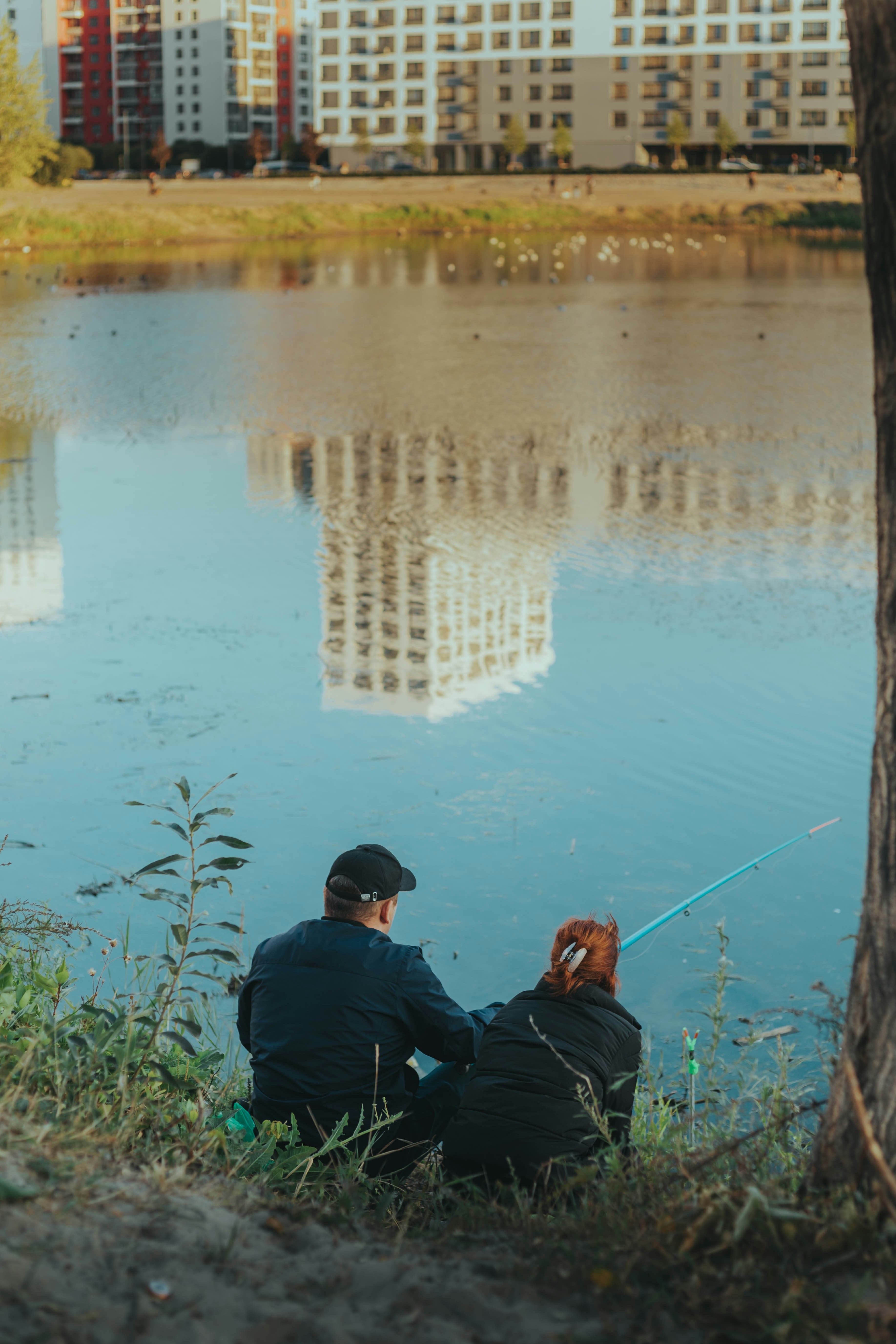 Two individuals fishing peacefully by a tranquil pond, with reflections of urban buildings shimmering on the water's surface.