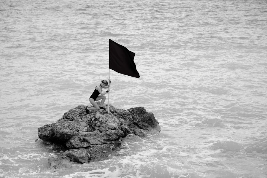 A person crouches on a small rock surrounded by water, holding a tall black flagpole. The monochrome scene feels stark and isolated.