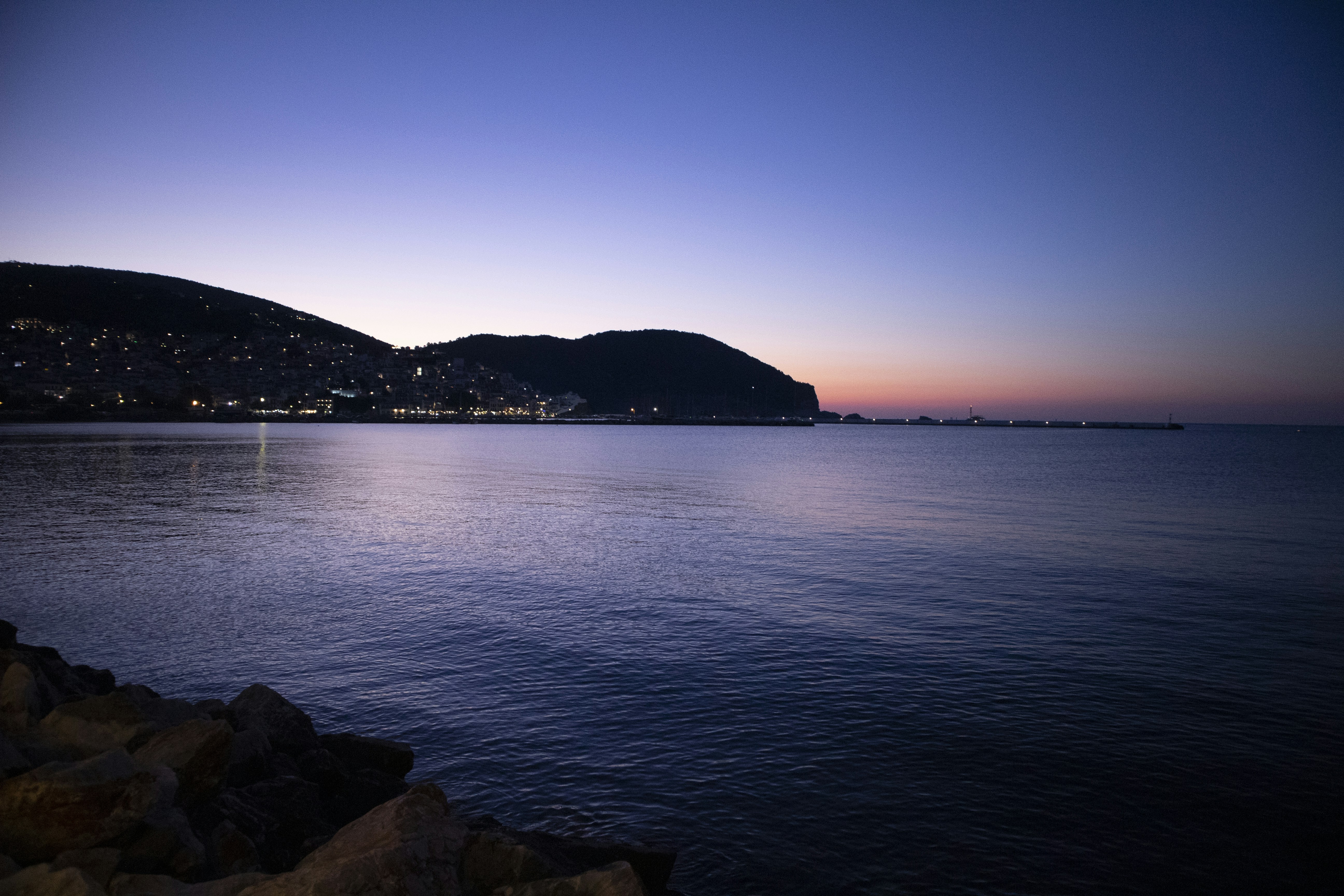 Gentle waves lapping against rocky shore under a twilight sky, with distant city lights shimmering on the water's surface.
