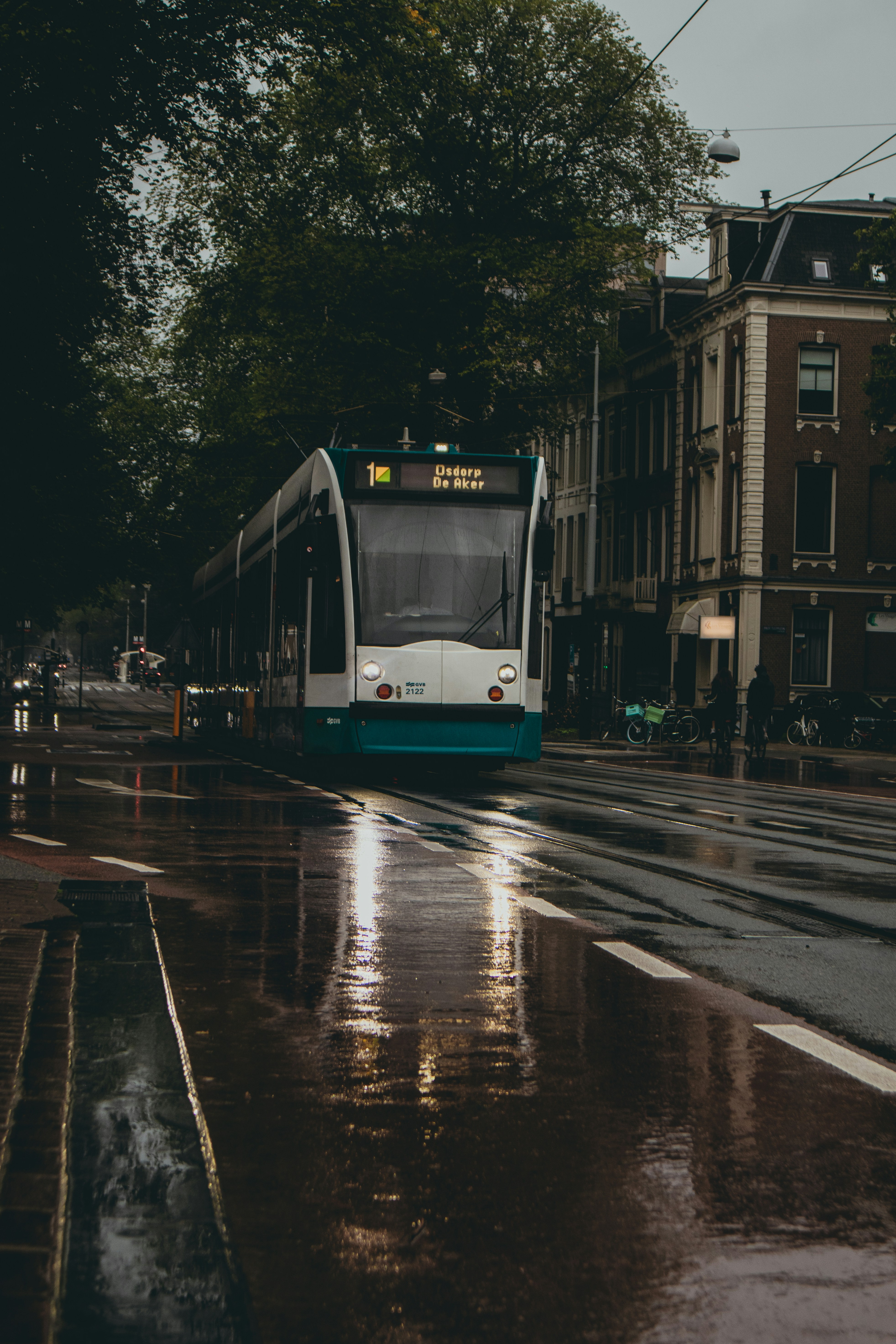 A modern tram glides through a rain-soaked street, reflecting the surrounding architecture and trees. The scene captures the essence of urban transit amidst a moody atmosphere.