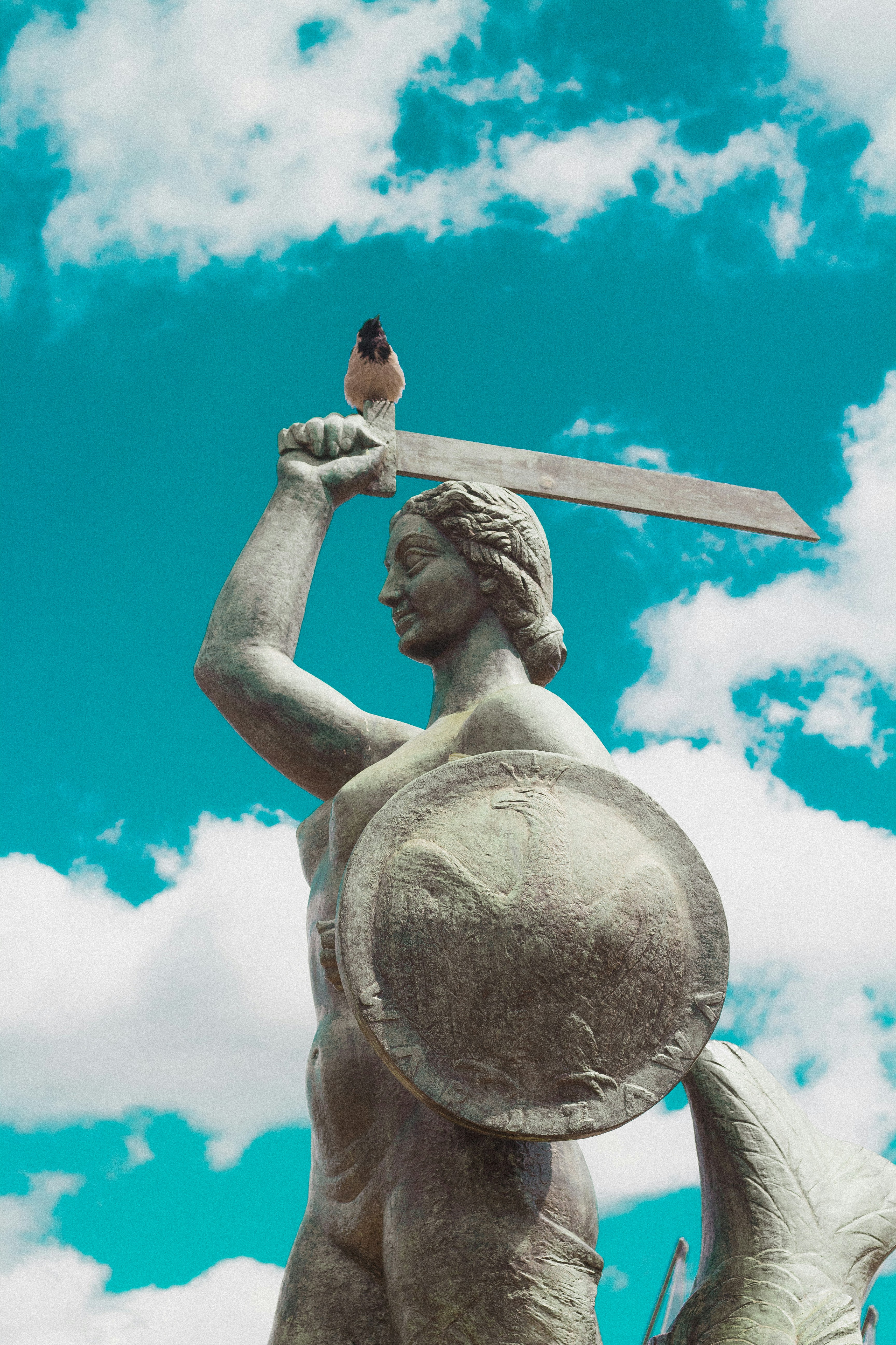 A bronze statue of a warrior holding a sword and shield, with a bird perched atop its sword against a bright blue sky dotted with clouds.