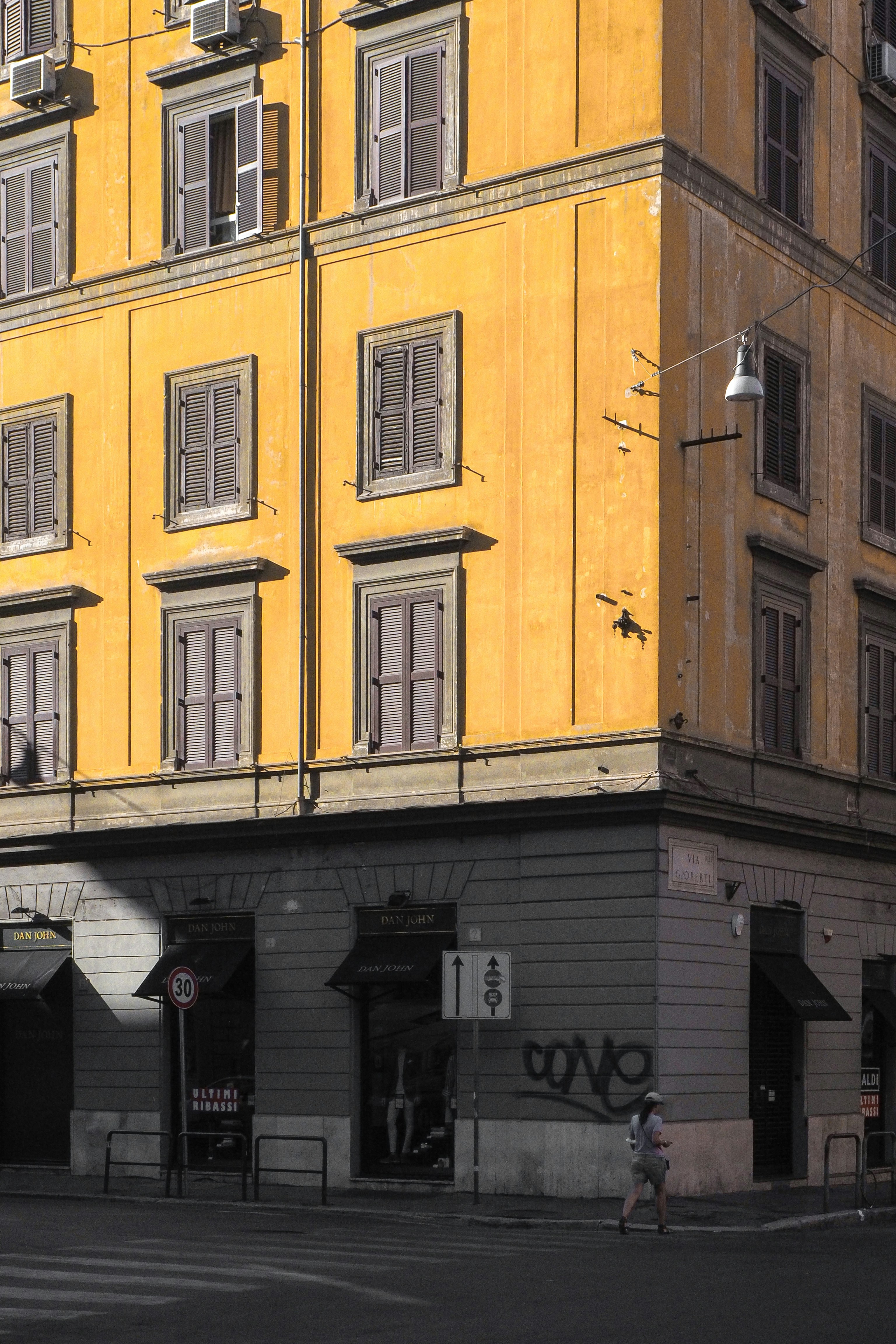 Bright yellow building corner with shuttered windows and street signage, showcasing urban architecture and everyday movement.
