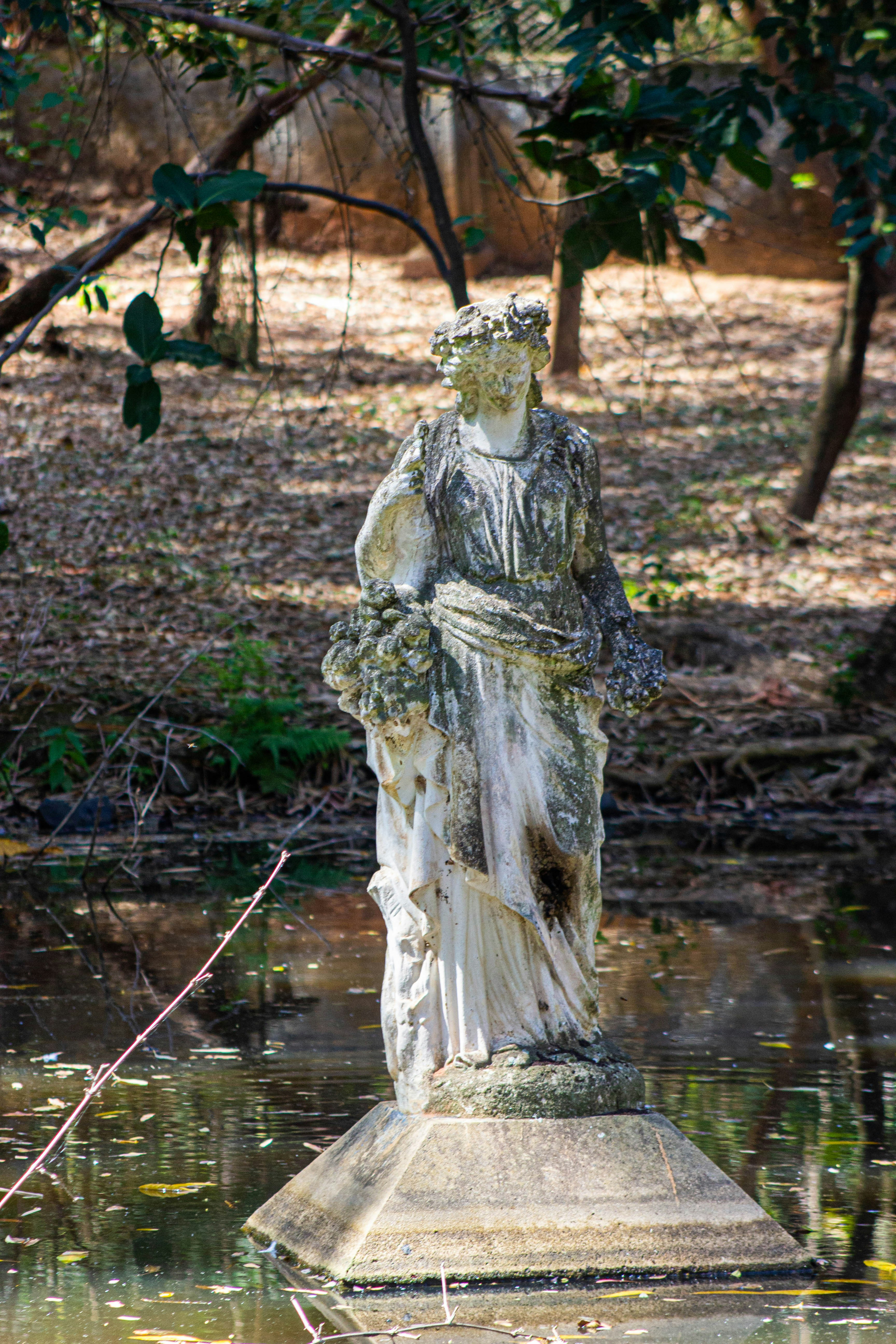 A timeworn statue of a woman adorned with floral elements stands solemnly on a pedestal in a tranquil pond, surrounded by lush greenery.