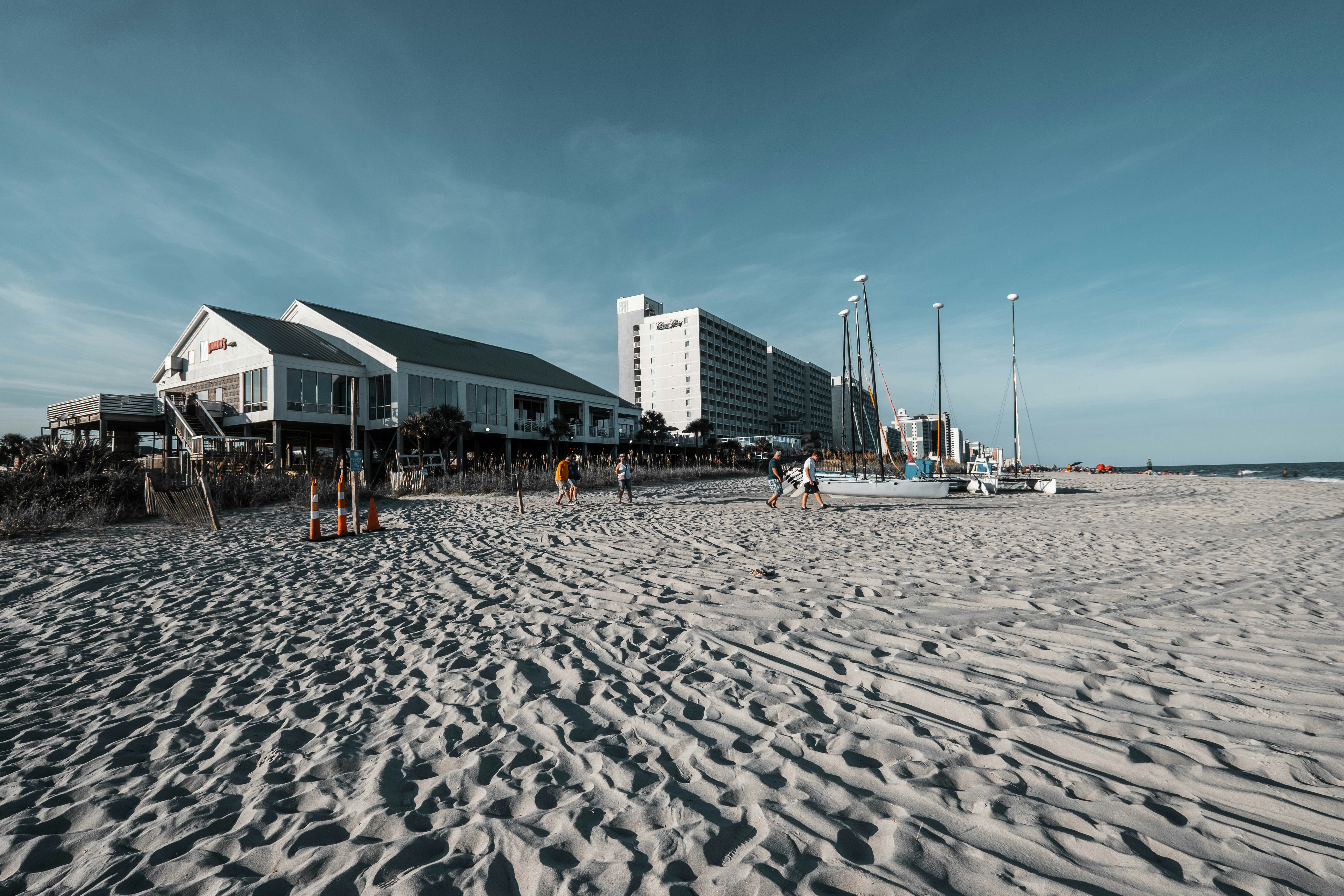 Beachgoers enjoying a sunny day on the sandy shore, with a beachside building and sailboats in the background.