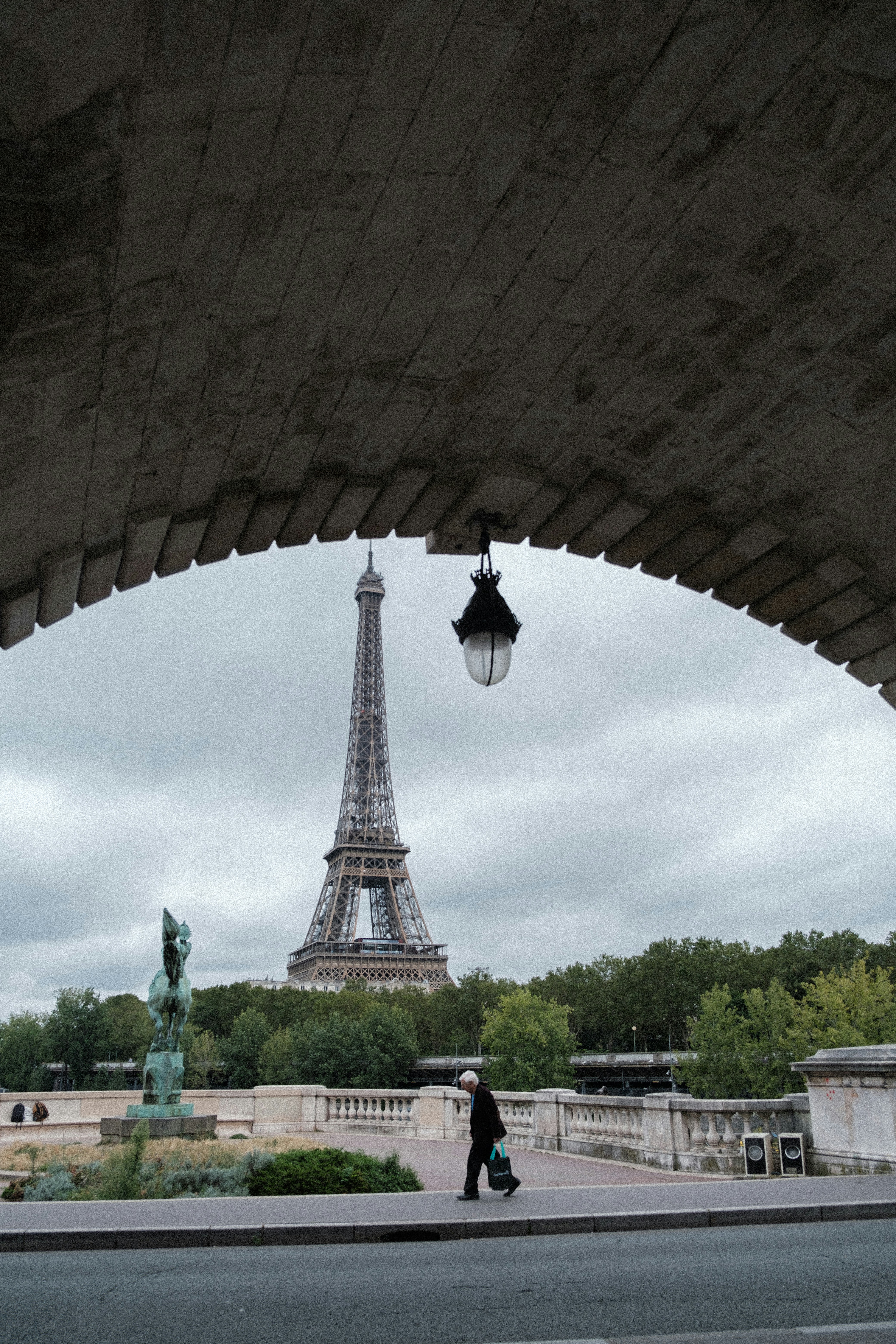 Eiffel Tower viewed through an archway, with a solitary figure walking along a tree-lined pathway. The scene encapsulates urban charm and historic architecture.