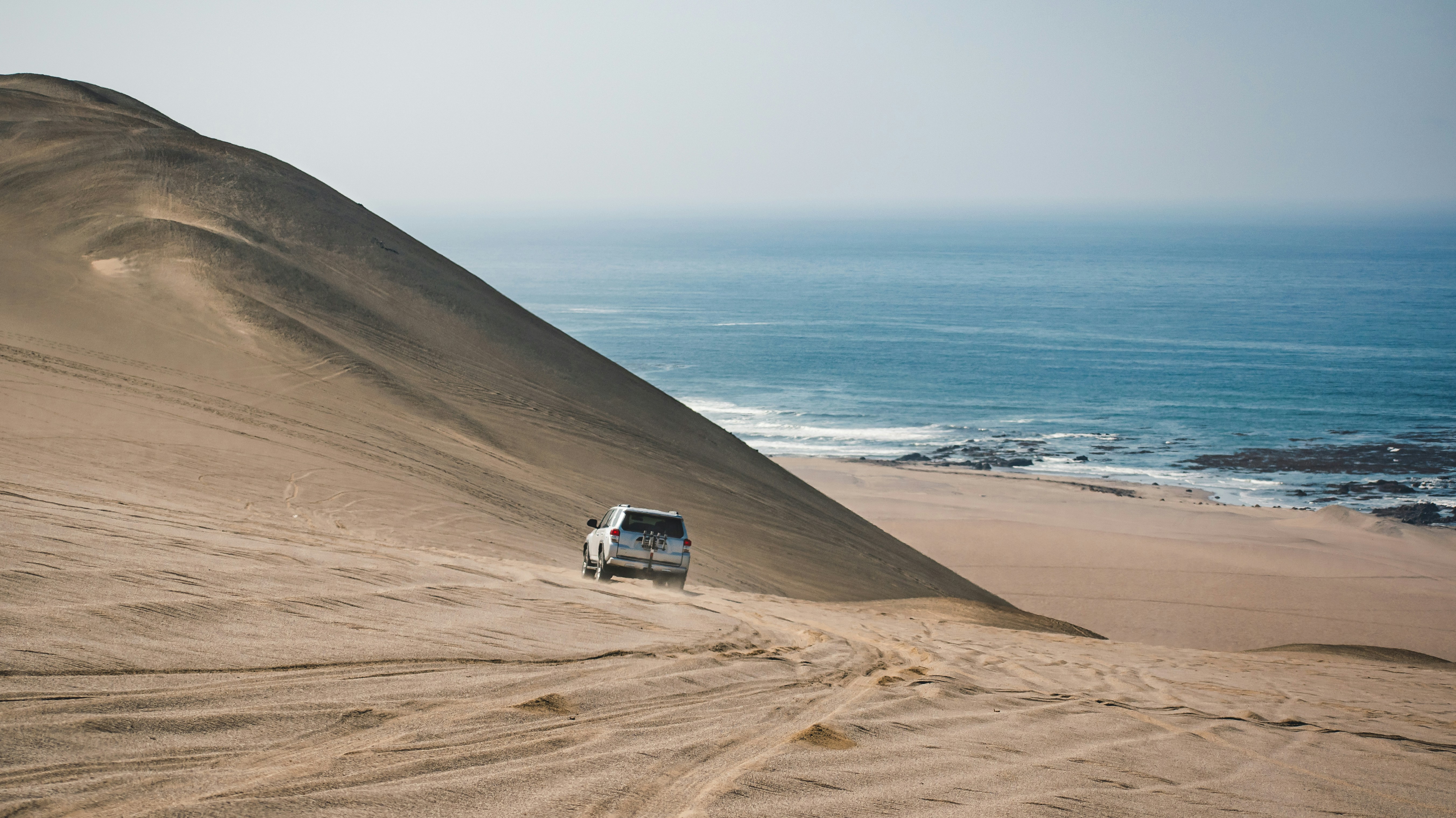 White SUV navigating a large sand dune with the ocean in the background.