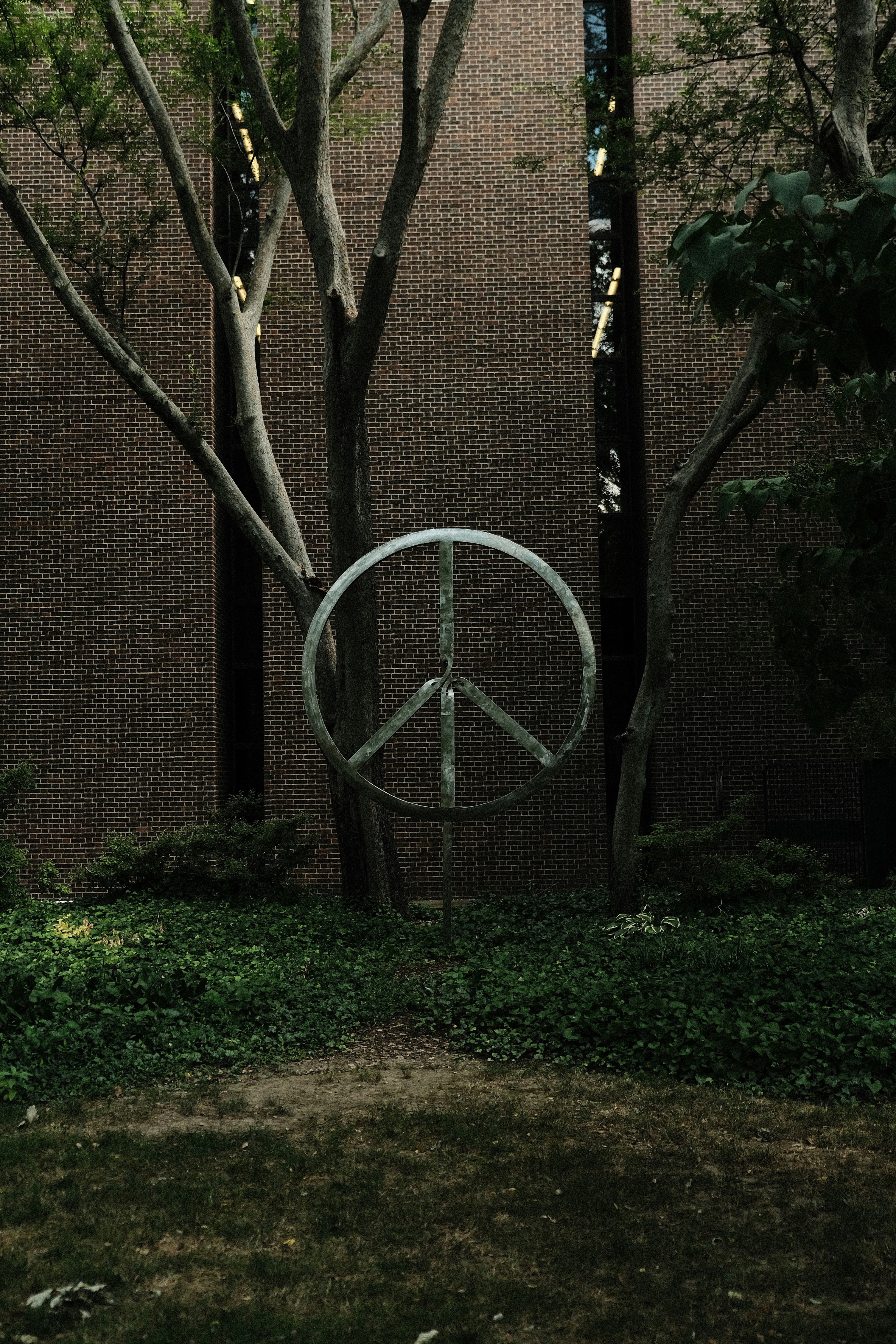 Metal peace symbol standing amidst trees and a brick wall, conveying a message of tranquility in an urban setting.