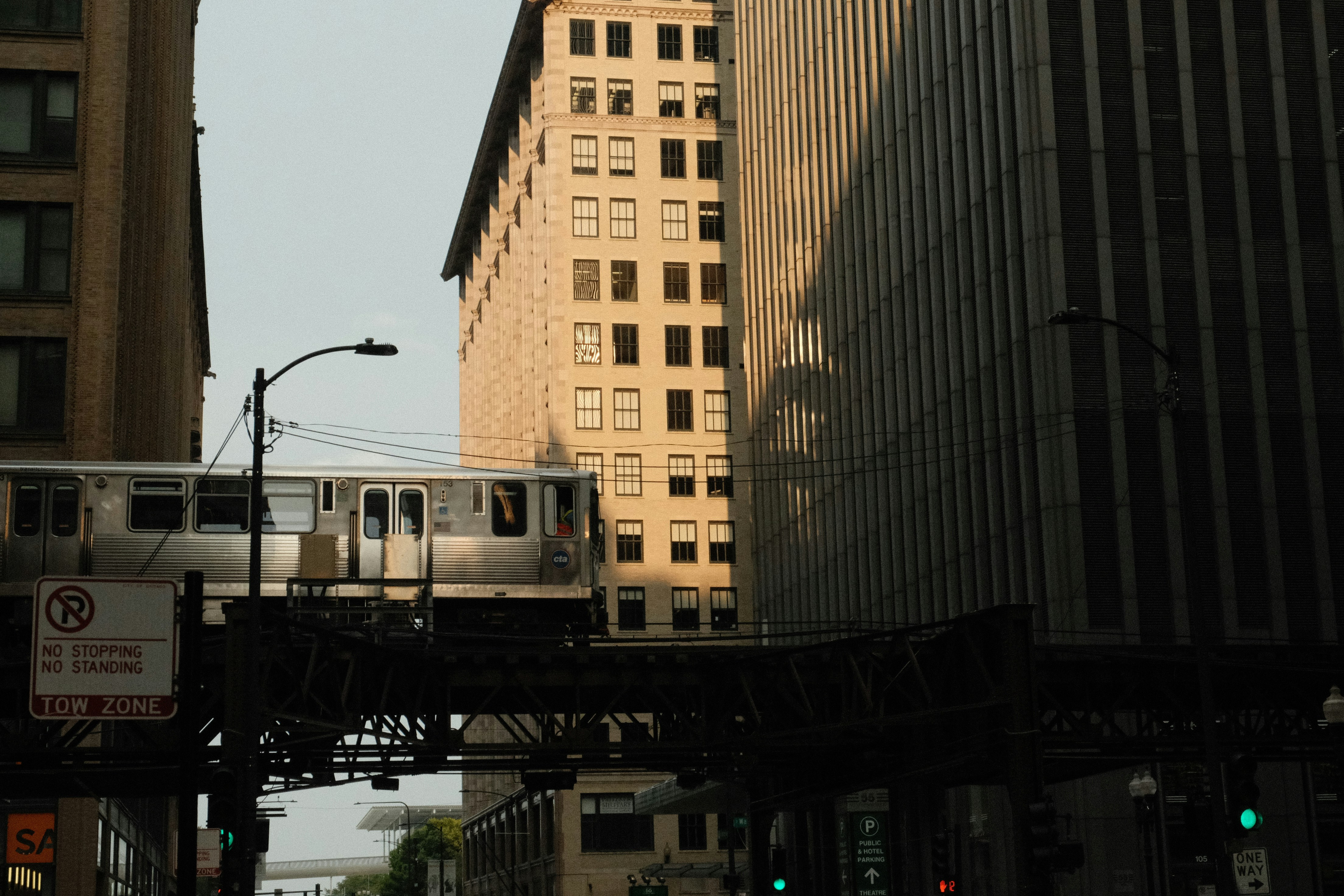 Silver train traversing elevated tracks against a backdrop of towering buildings, casting intricate shadows on the urban landscape.