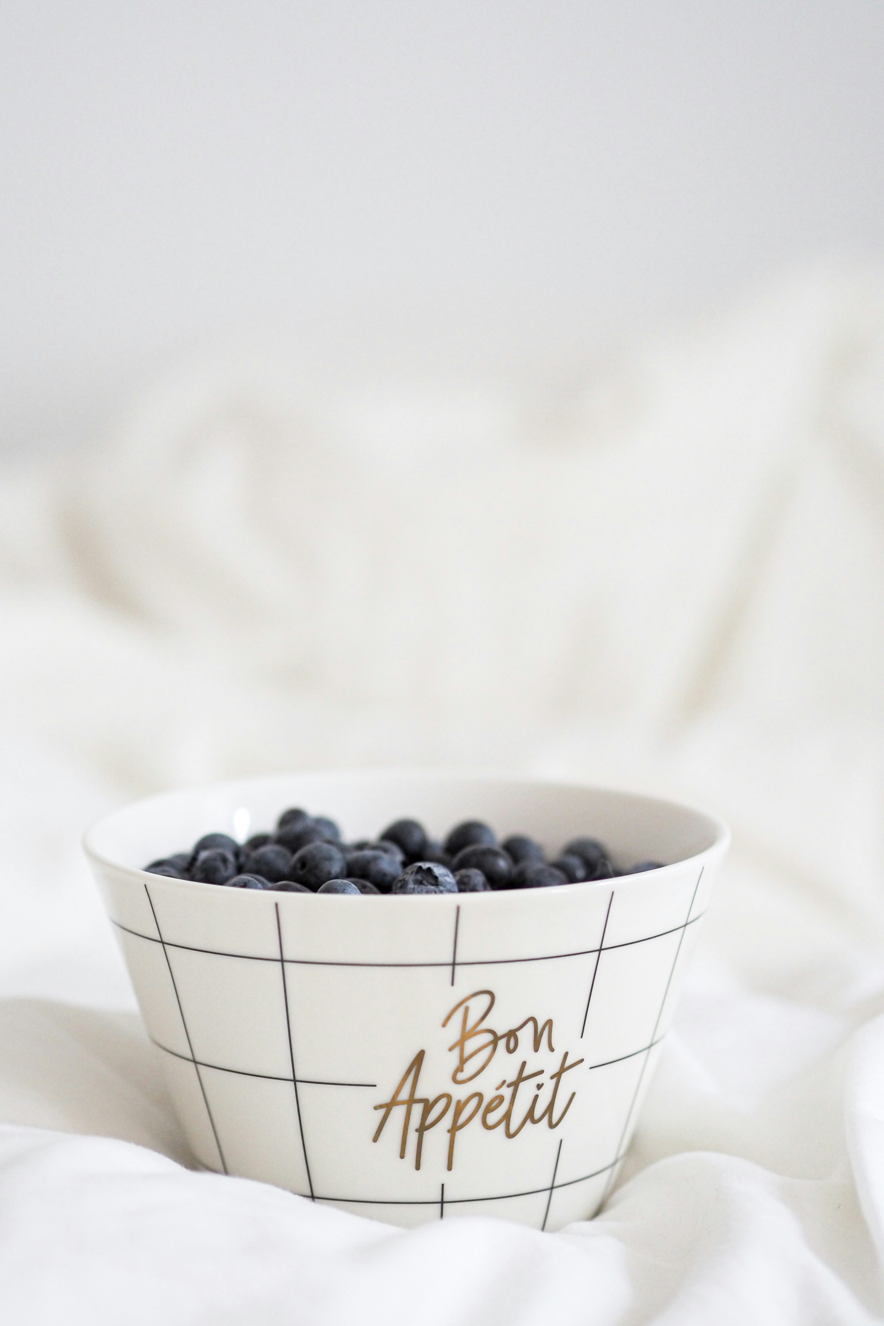White bowl filled with fresh blueberries, adorned with 'Bon Appétit' inscription, set against a soft, neutral background.