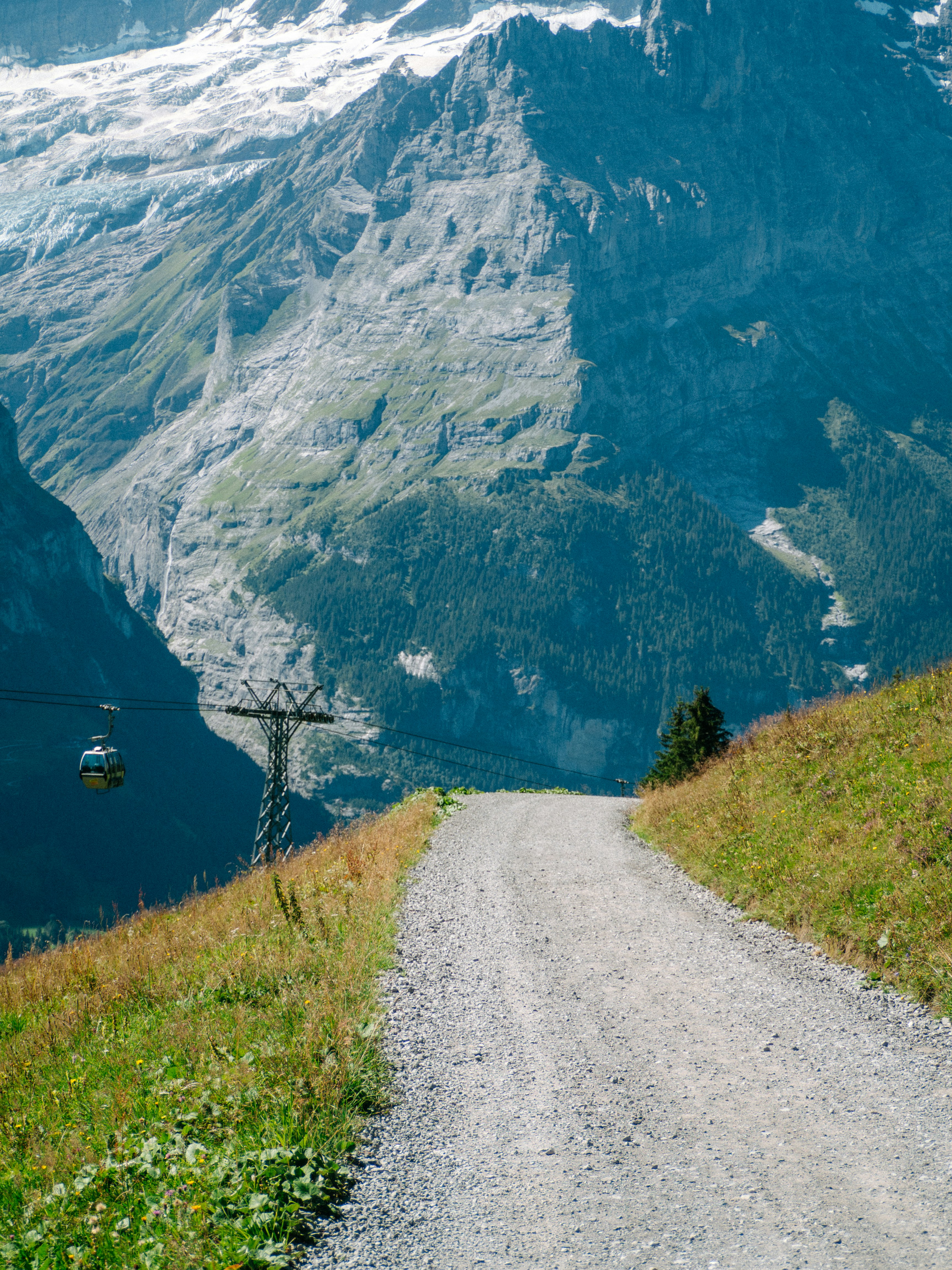 Gravel pathway leading towards towering mountains under a clear blue sky, with a gondola traversing the landscape.