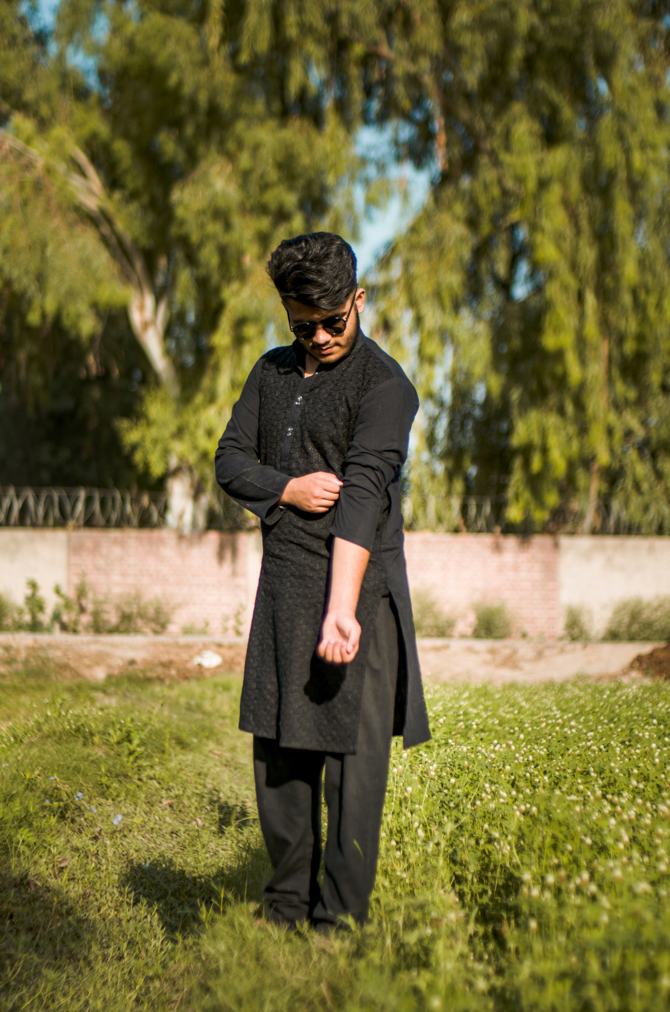 Man in a black kurta stands in a sunlit meadow, adjusting his sleeve as he looks downward. Tall trees and a brick wall form a calm, natural backdrop for a straightforward portrait.