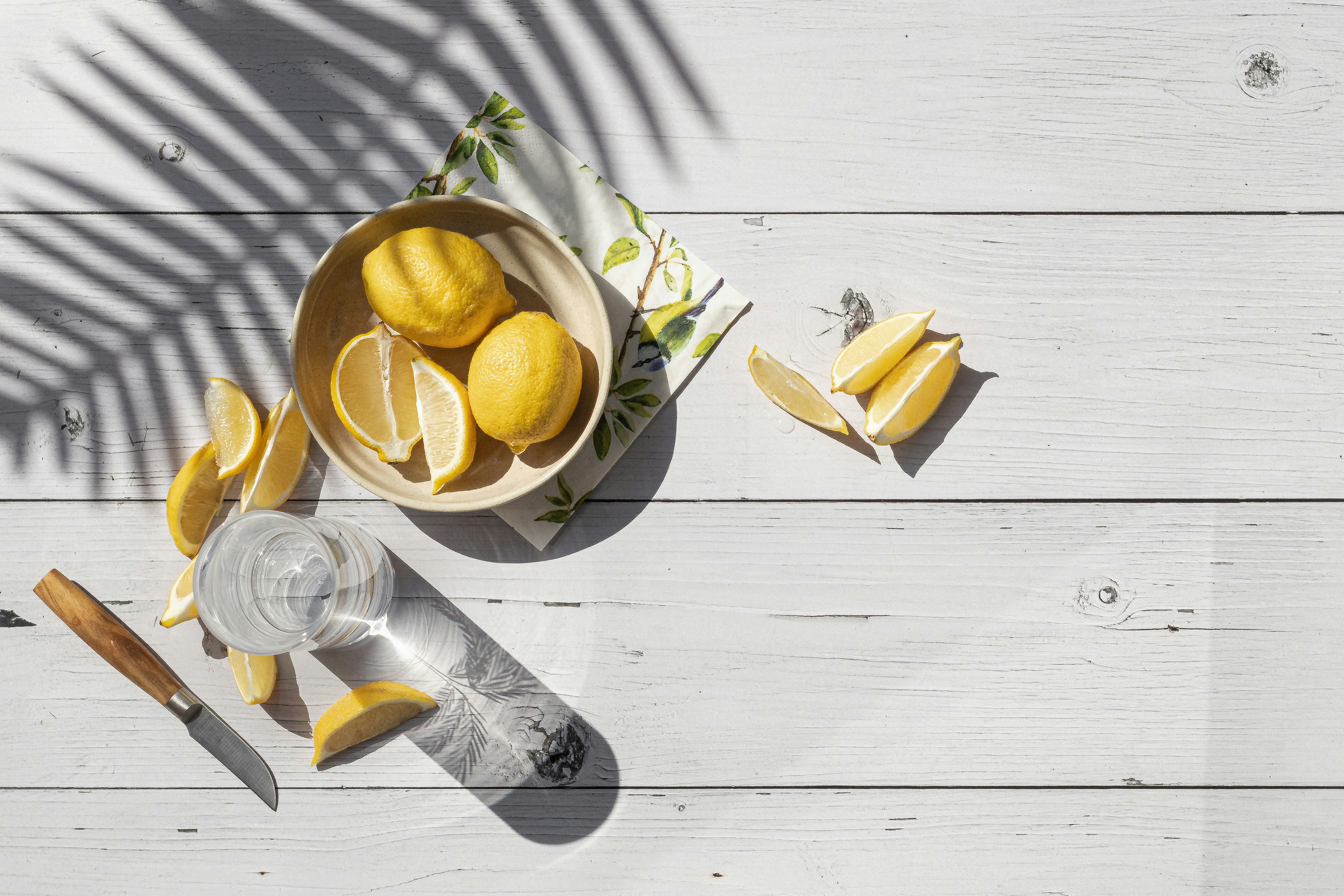 Lemons in a bowl with sliced pieces and a knife on a white wooden surface, accented by palm shadows.