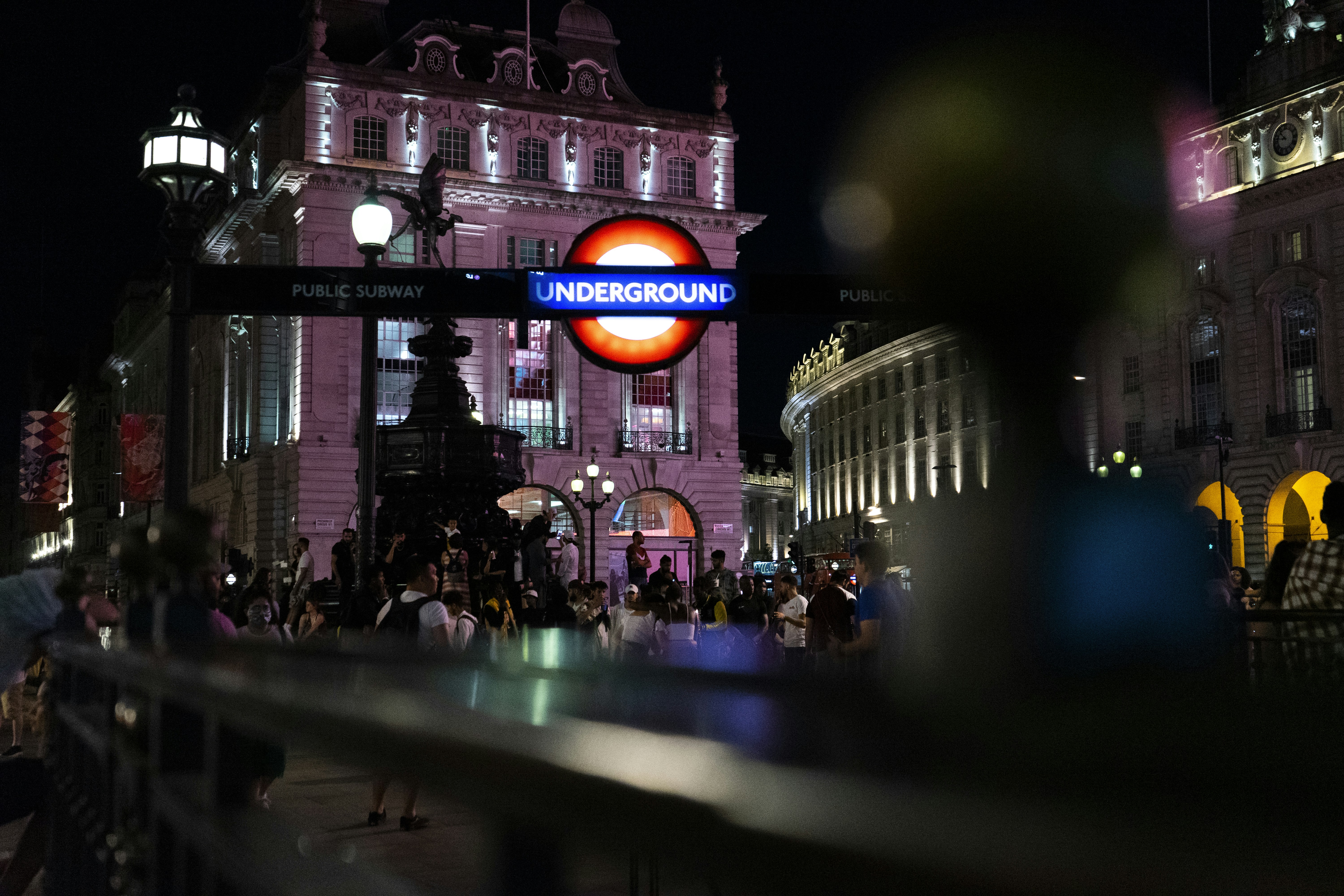 Night photograph of a London Underground entrance with a glowing roundel sign amid a crowd. Railings in the foreground frame the scene while a historic building lights up in the background.