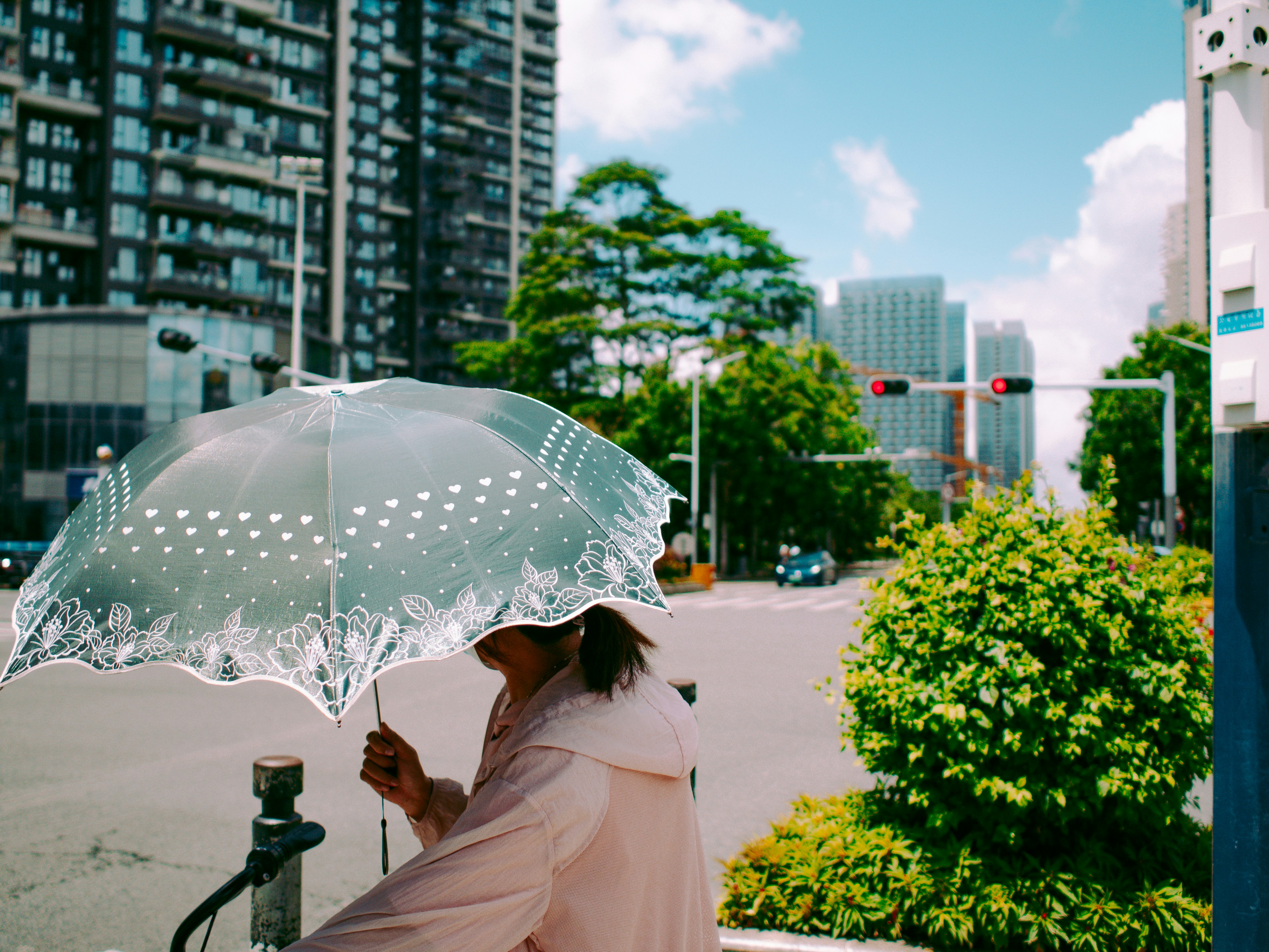 A woman with a lace umbrella walks down a sunlit city street. Modern high-rises and trimmed shrubs line the sidewalk.