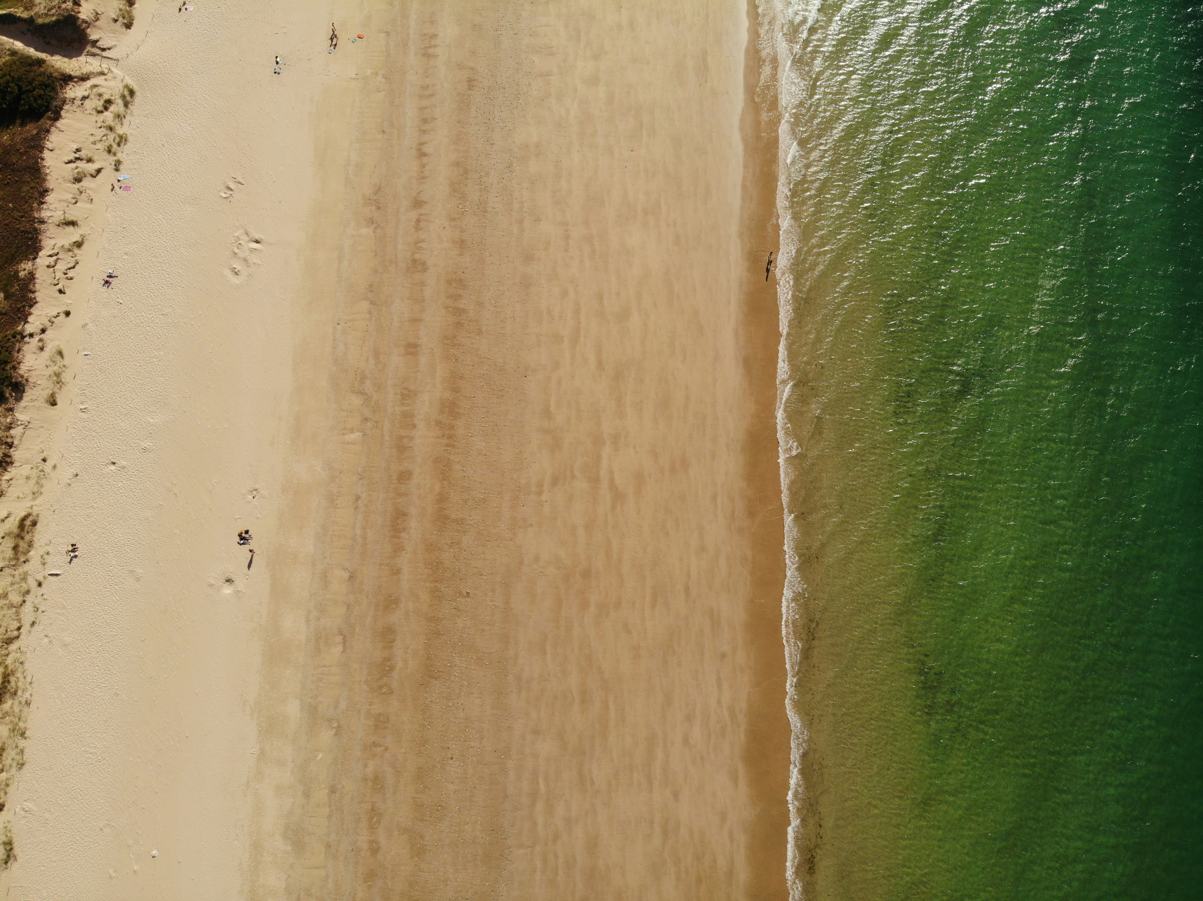 Aerial view of a sandy beach transitioning into emerald waters, showcasing footprints and gentle waves. The natural gradient of colors enhances the serene coastal landscape.