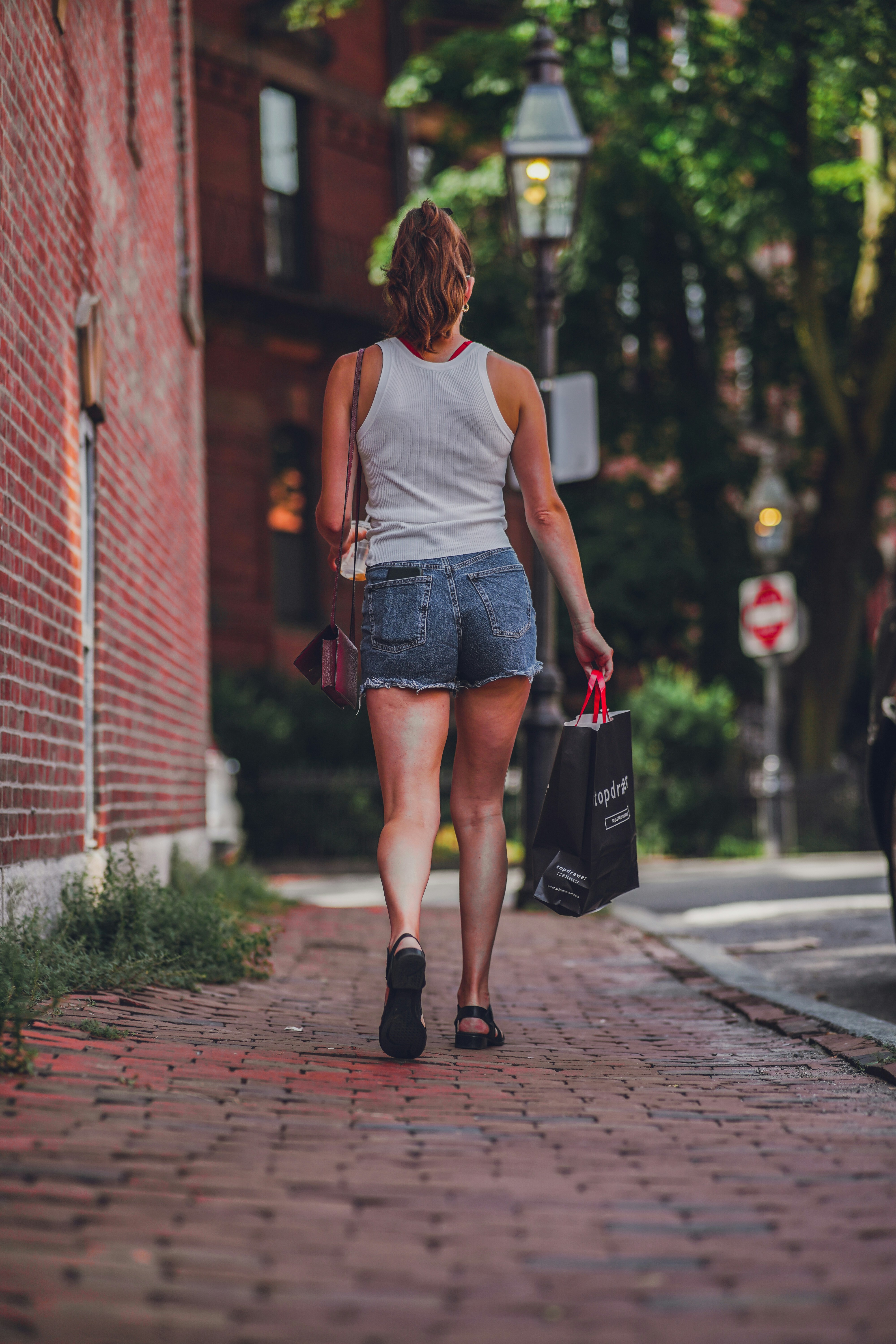 Back-Profile Stroll: Denim Shorts and Shopping Bags on a Brick City Walk