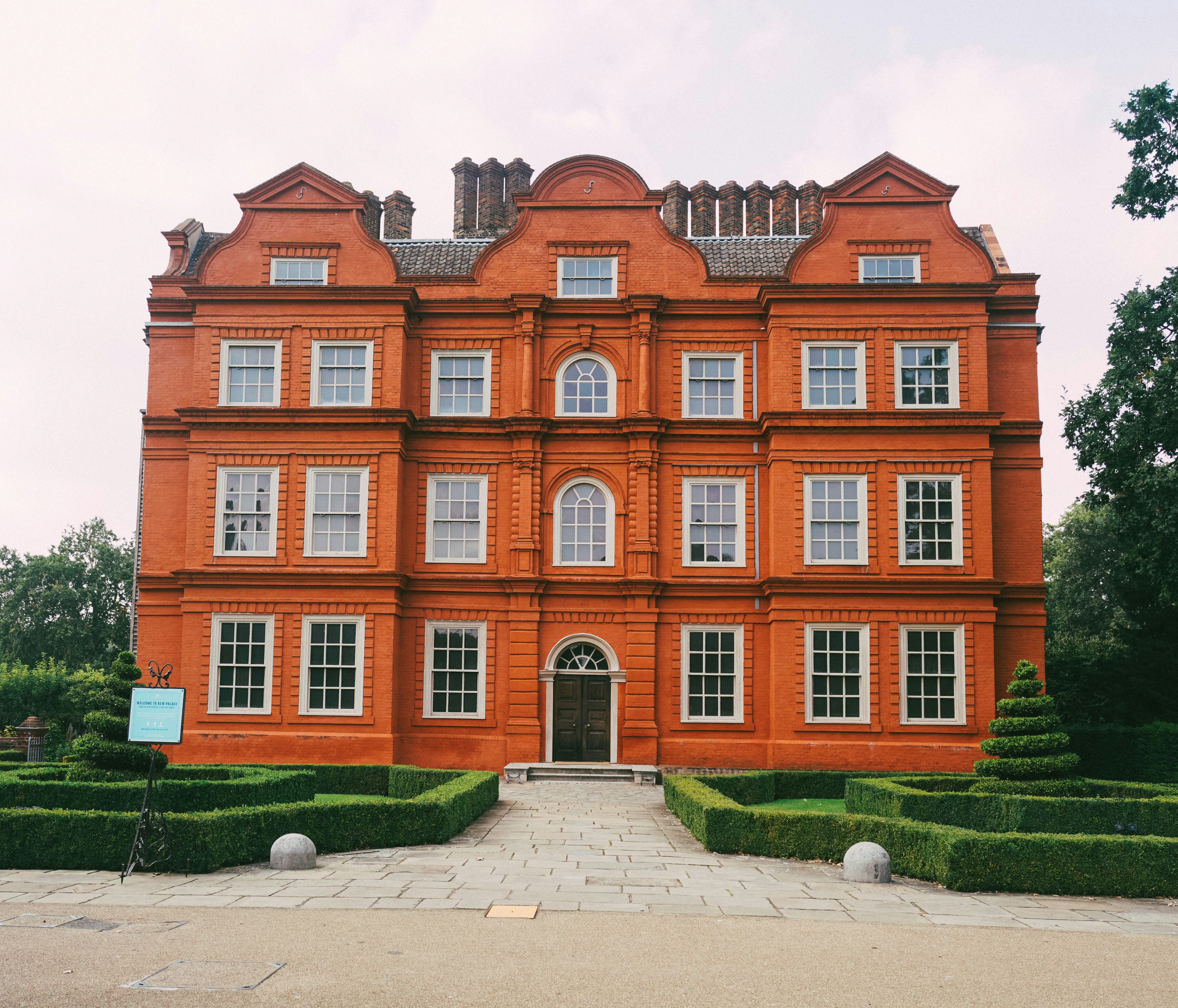 This striking image captures a grand red brick mansion, showcasing its symmetrical architecture and classic Georgian style. The building's vivid orange-red hue stands out against the soft greenery surrounding it, while the clear blue sky adds a serene atmosphere. The composition is perfectly balanced, drawing the viewer's eye to the intricate details and vibrant colors that make the mansion a captivating focal point.