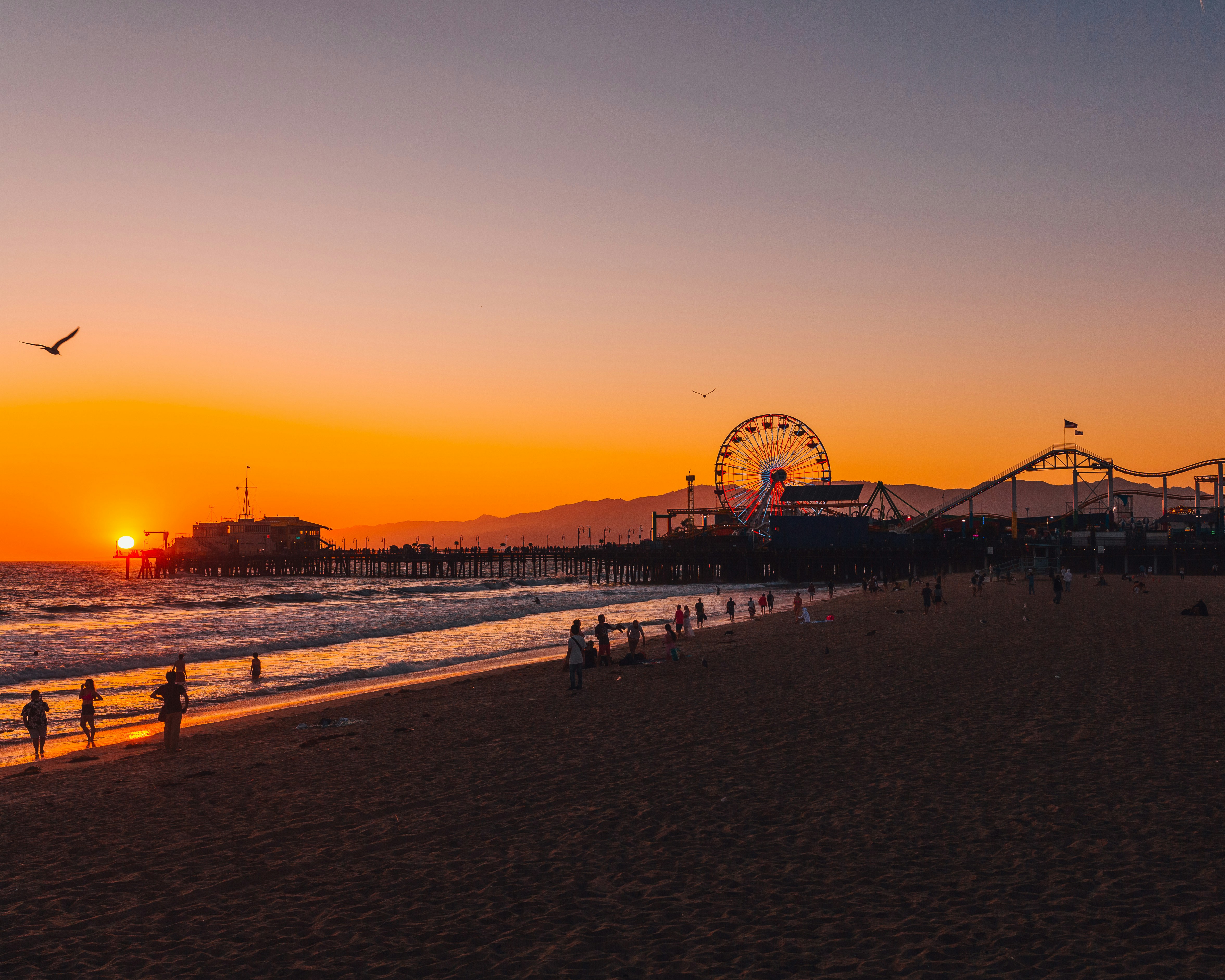 Silhouetted figures stroll along the sandy beach as the sun sets behind a vibrant amusement pier, with a ferris wheel illuminated against the twilight sky.