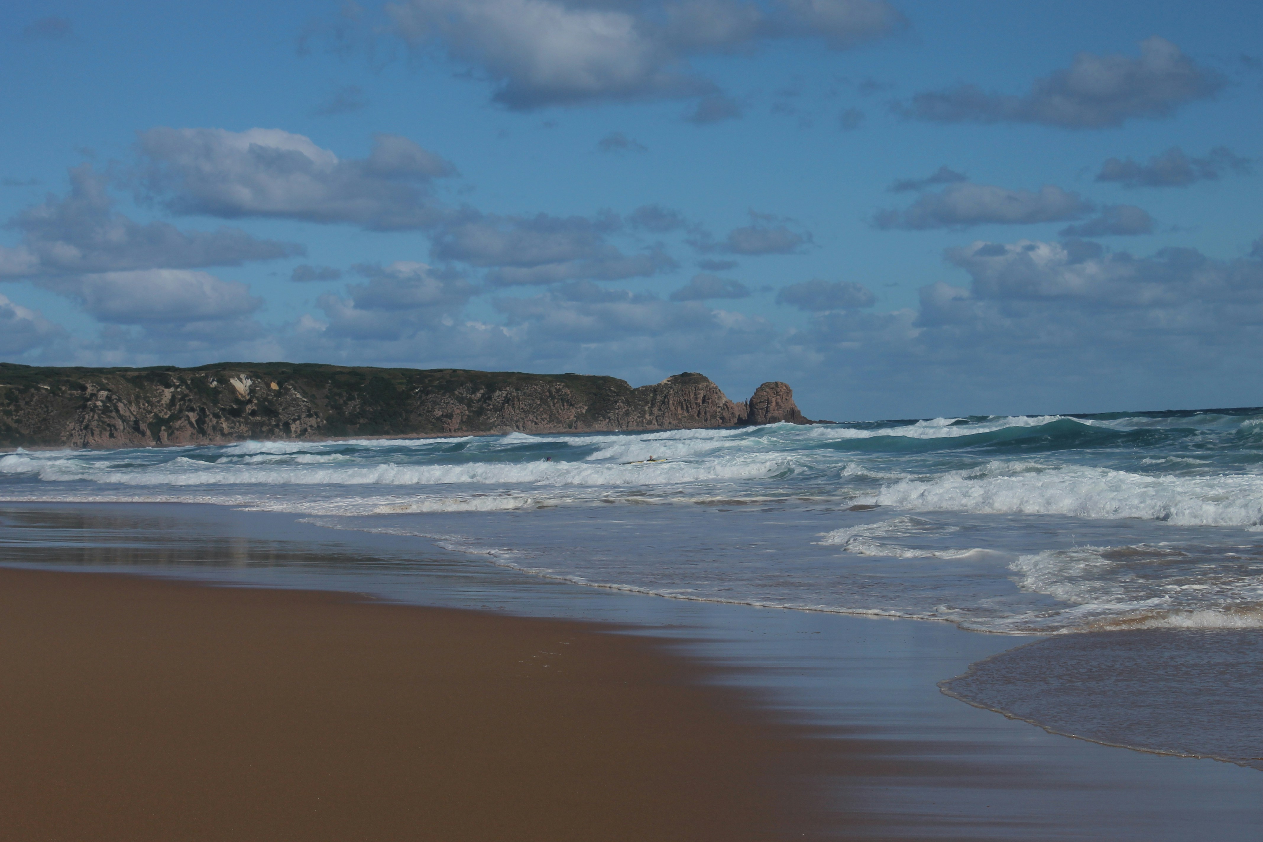Waves gently lap against a sandy beach, with rocky cliffs rising in the background under a partly cloudy sky.