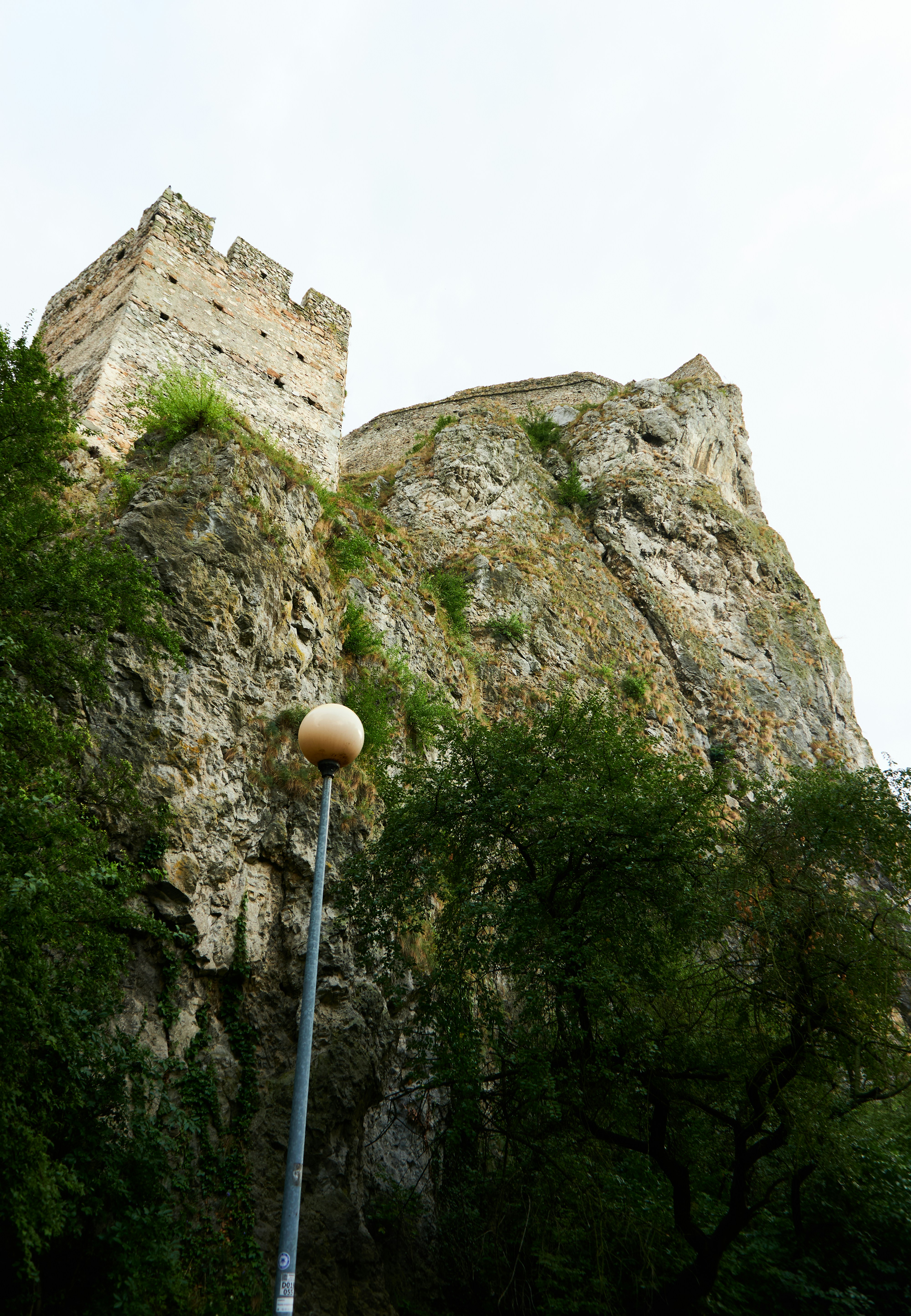 Ancient stone fortress perched atop a rugged cliff, surrounded by lush greenery and a cloudy sky.