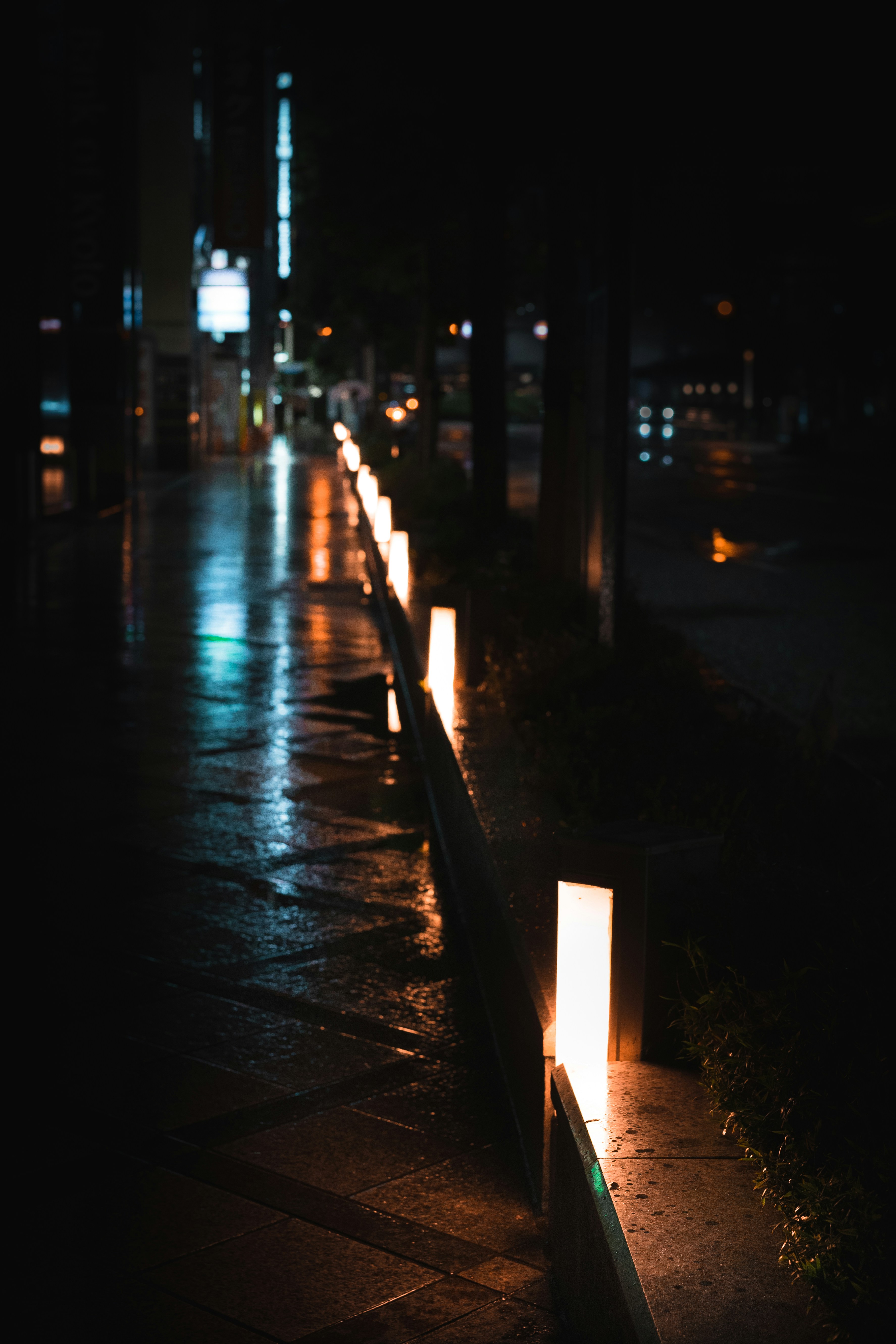 Wet pavement glistens under glowing light fixtures along a city sidewalk at night.