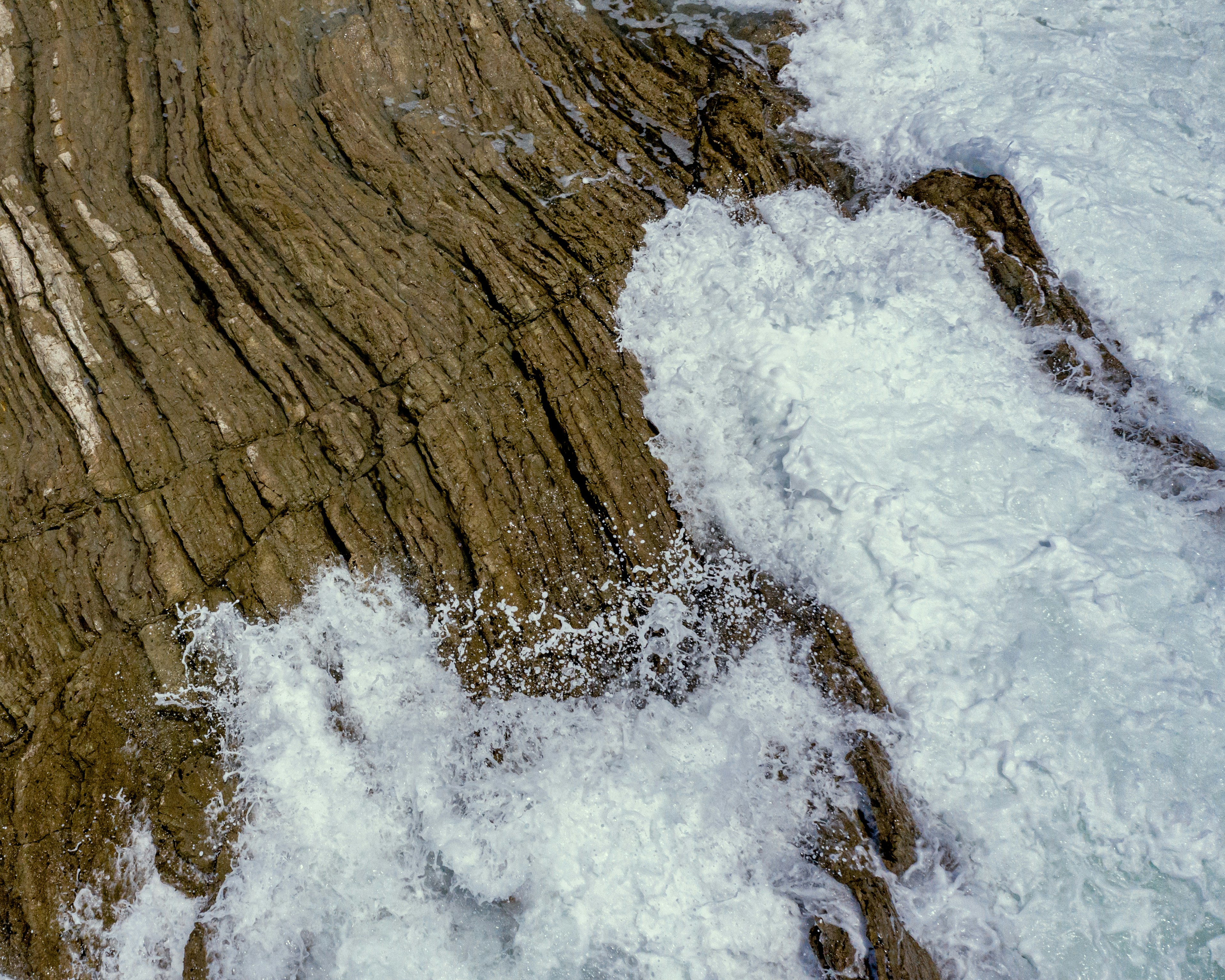 Rugged rock formations meet frothy waves, showcasing the dynamic interplay between land and sea. The natural patterns etched into the stone create a striking contrast with the flowing water.