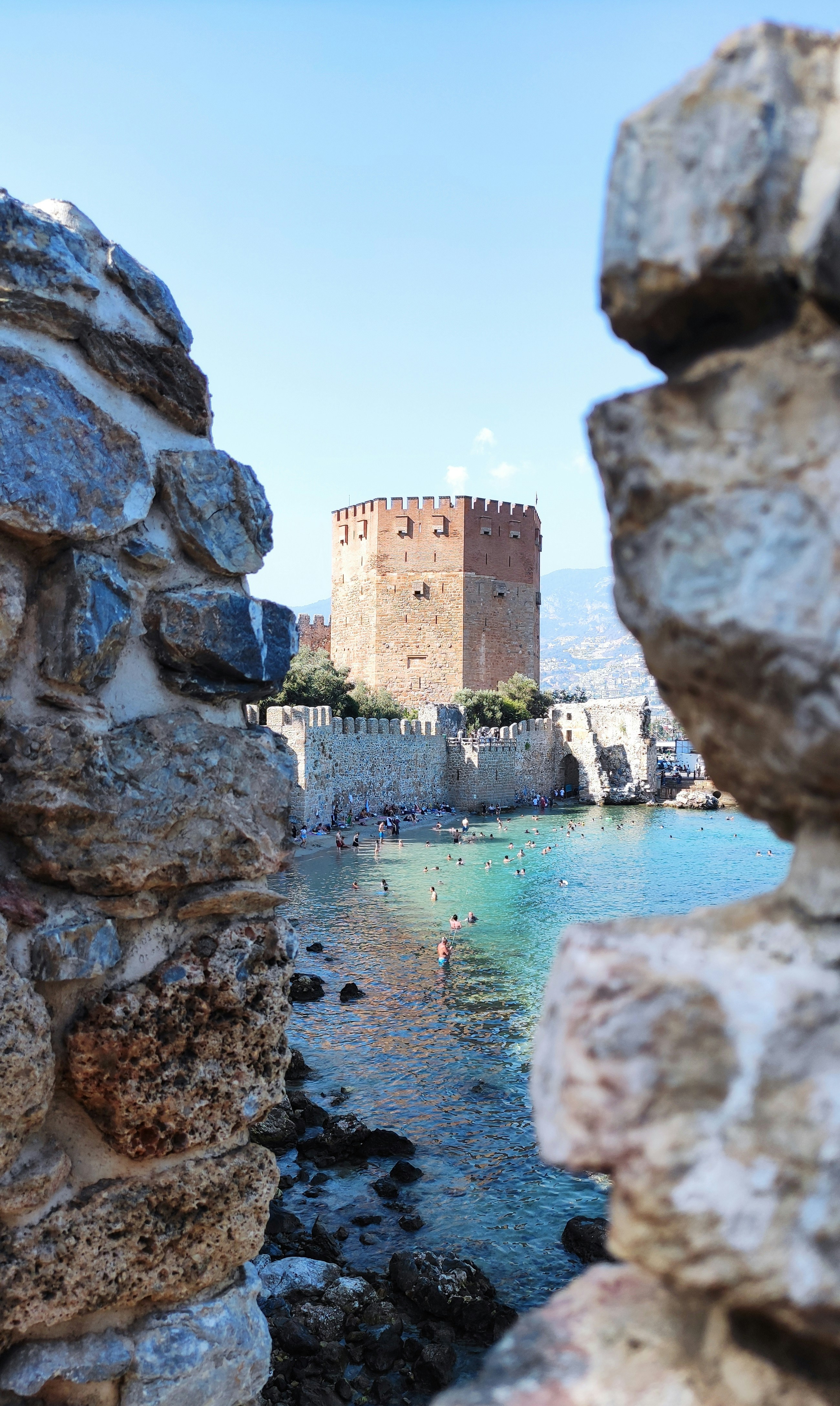 A historic tower framed by ancient stones, overlooking a vibrant beach filled with sunbathers and swimmers. The turquoise waters reflect the clear sky.