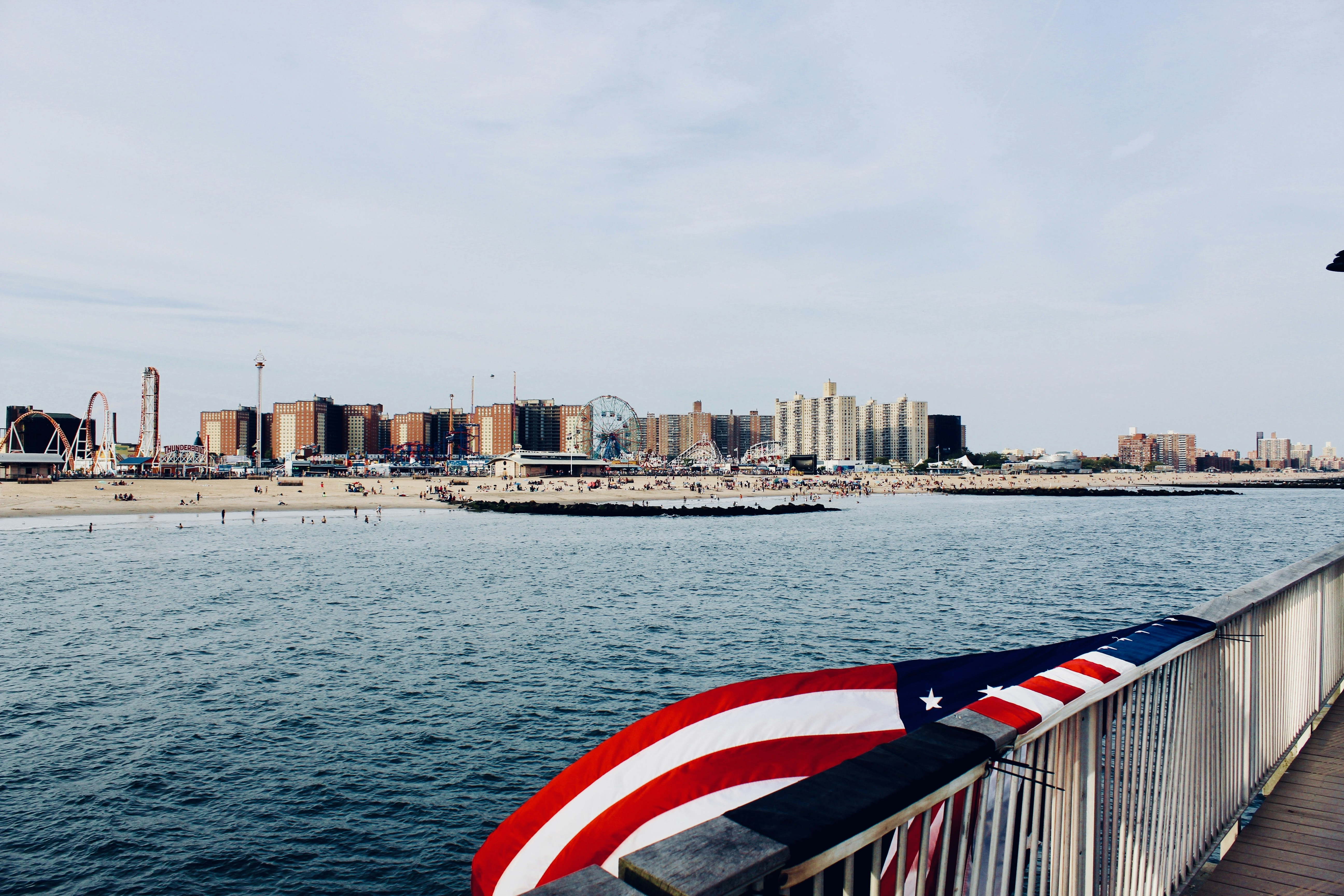 American flag waving gently on a pier, overlooking a lively beach scene with amusement rides and city buildings in the background.