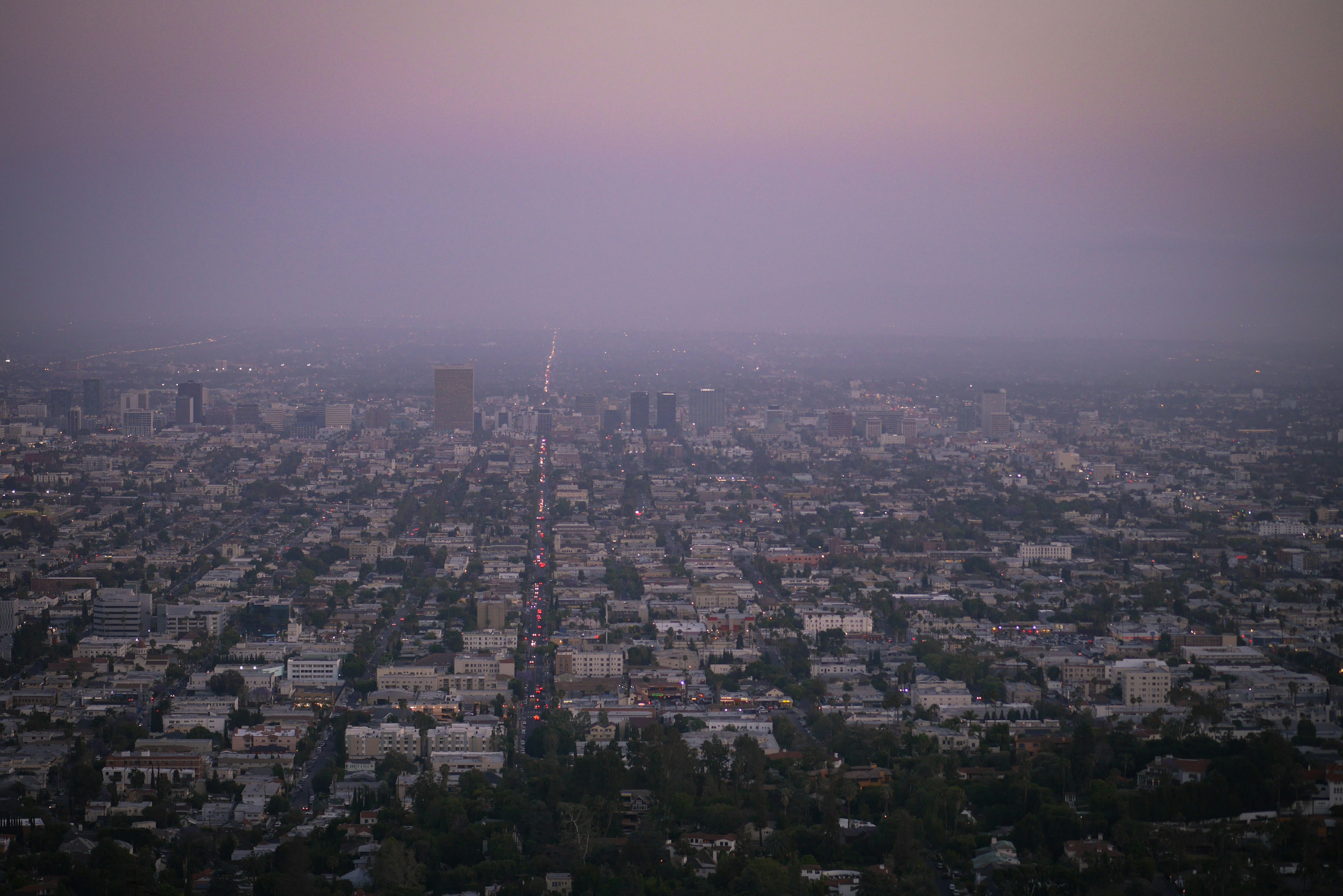 Aerial view of a sprawling cityscape at twilight, showcasing a grid of buildings under a hazy sky with faint streetlights illuminating the main thoroughfare.