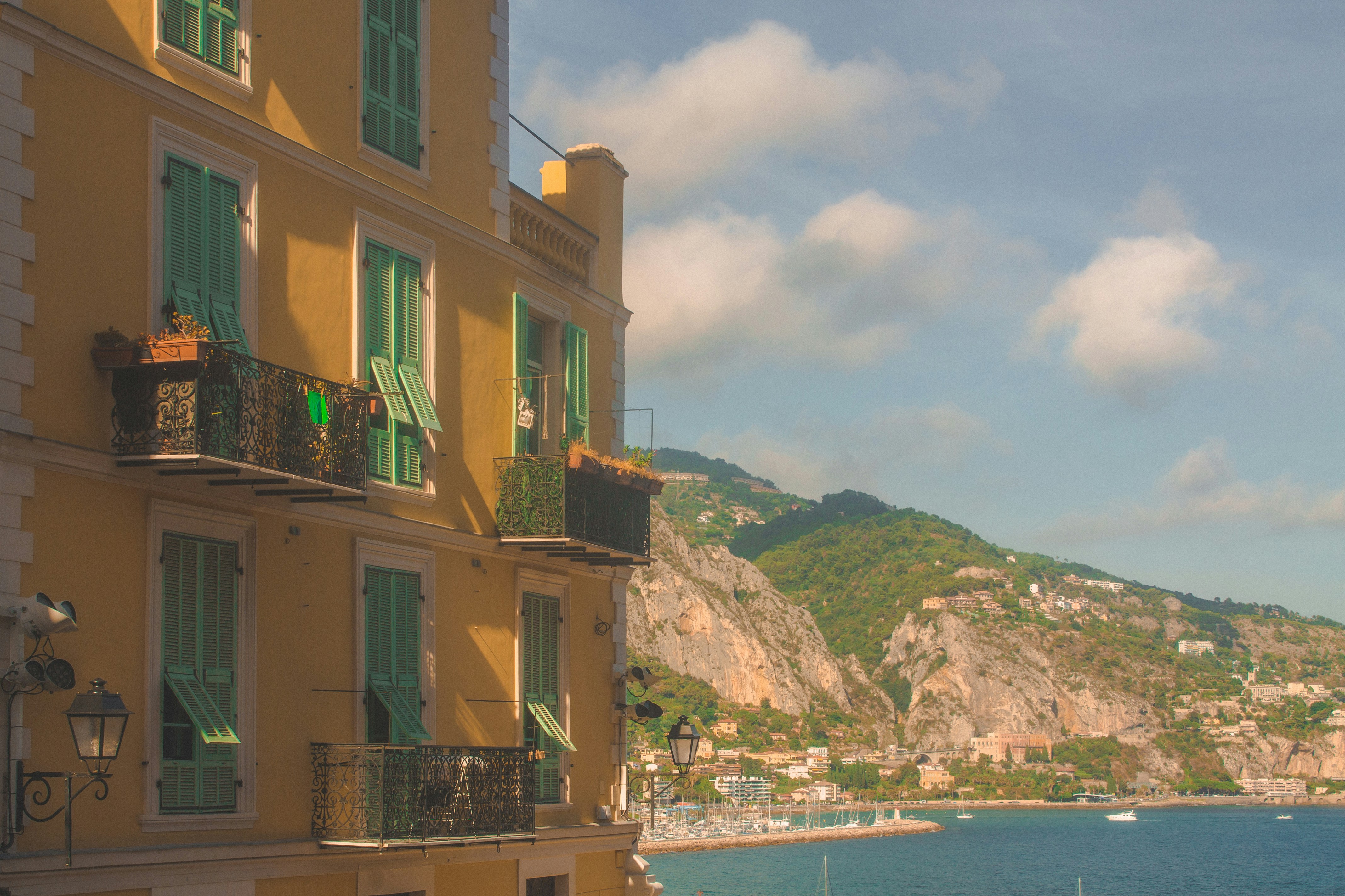 Yellow building with green shutters and balconies facing a mountainous coastline under a partly cloudy sky.