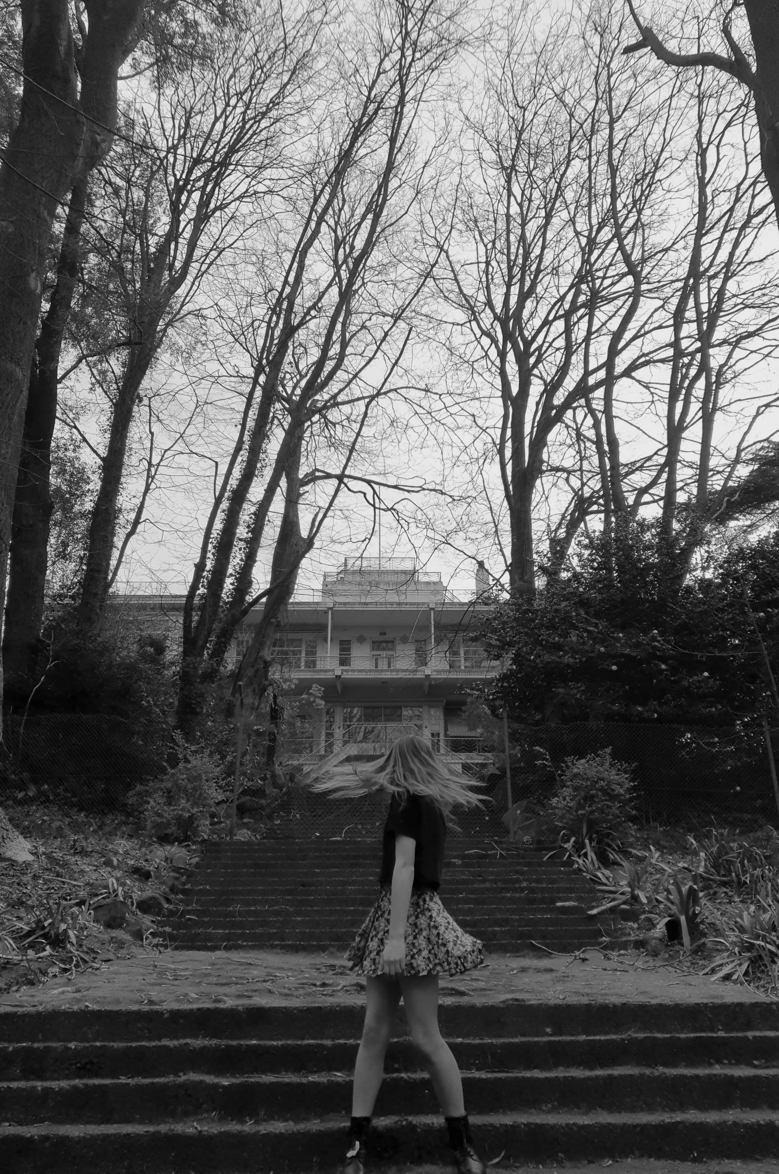A figure in a swirling skirt stands on worn steps, framed by stark, leafless trees and an abandoned building in the background.