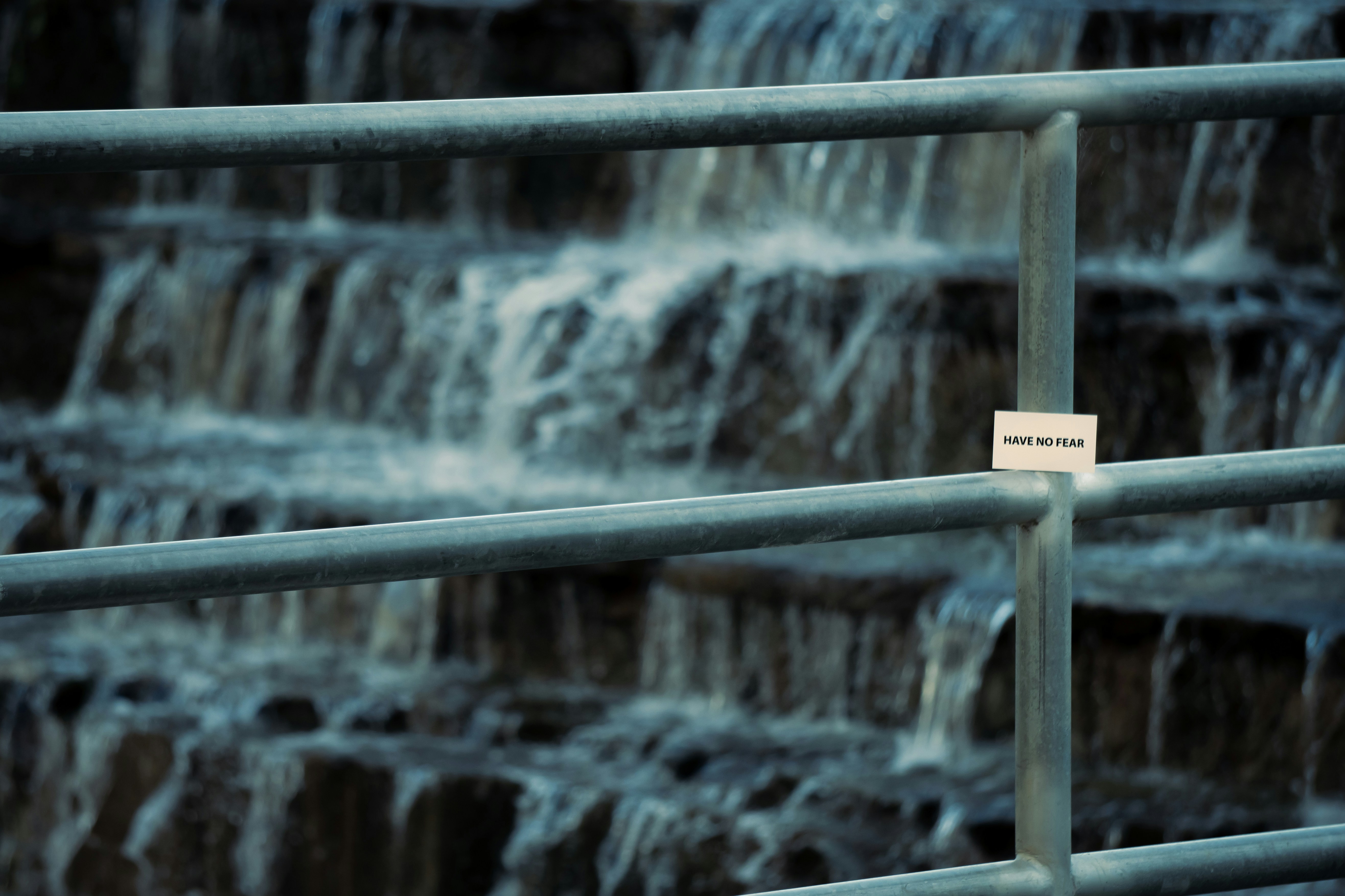 A tranquil waterfall cascades over rocky tiers, with a small sign reading 'HAVE NO FEAR' resting on a railing nearby.