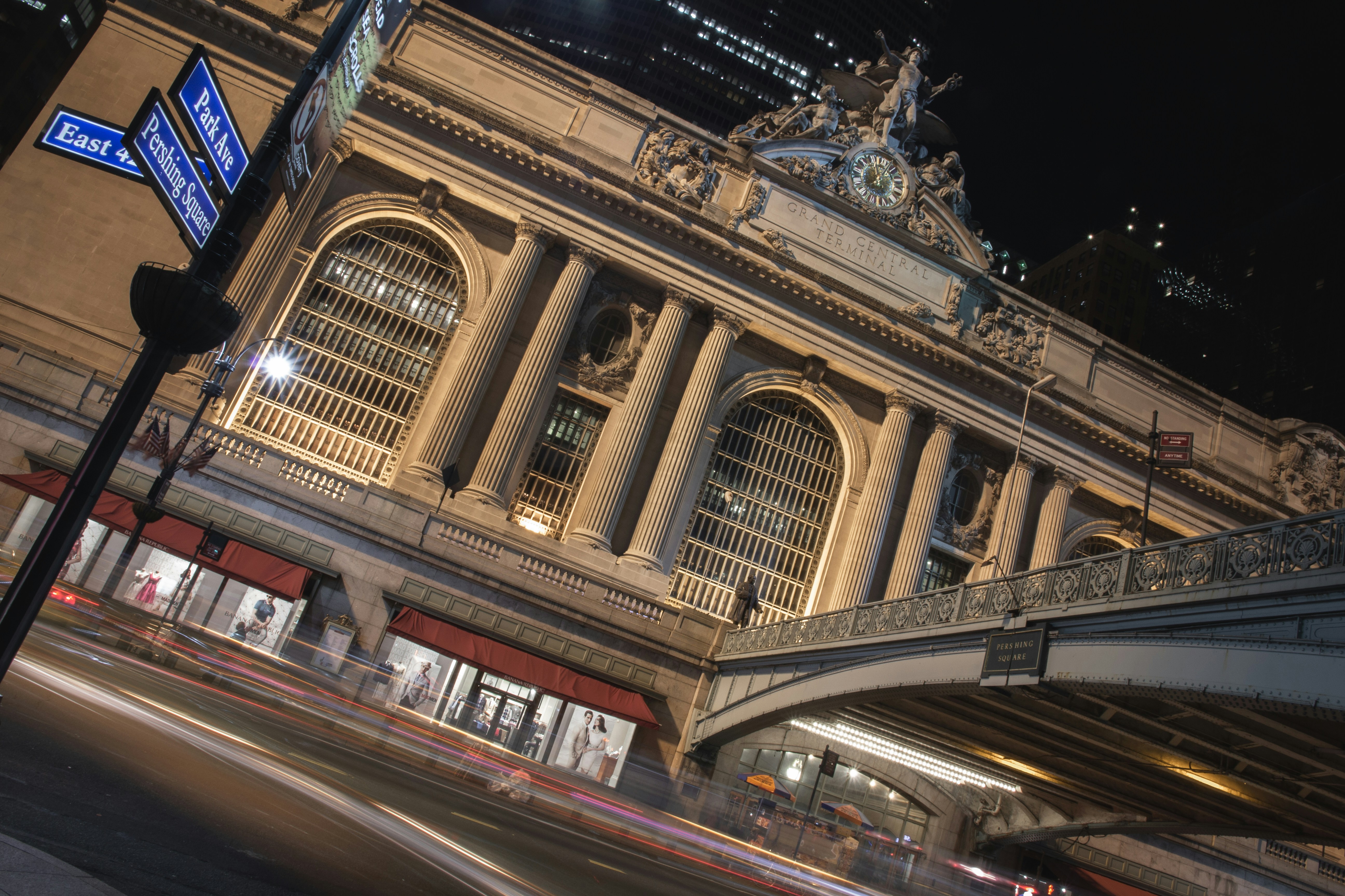 Facade of a grand historic station framed by a curved bridge and illuminated street