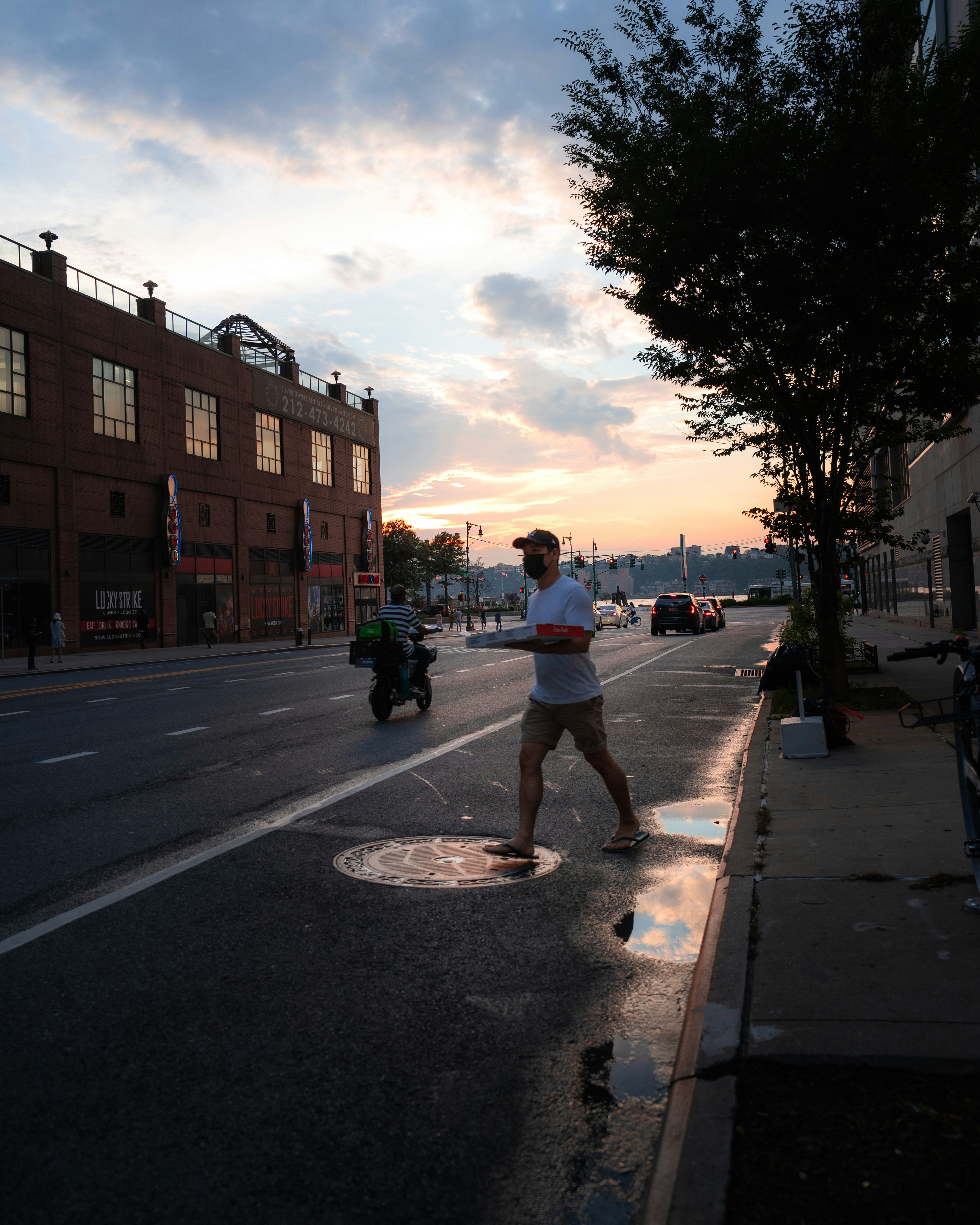 Delivery person walking along a wet street at sunset, with urban buildings and vehicles in the background.